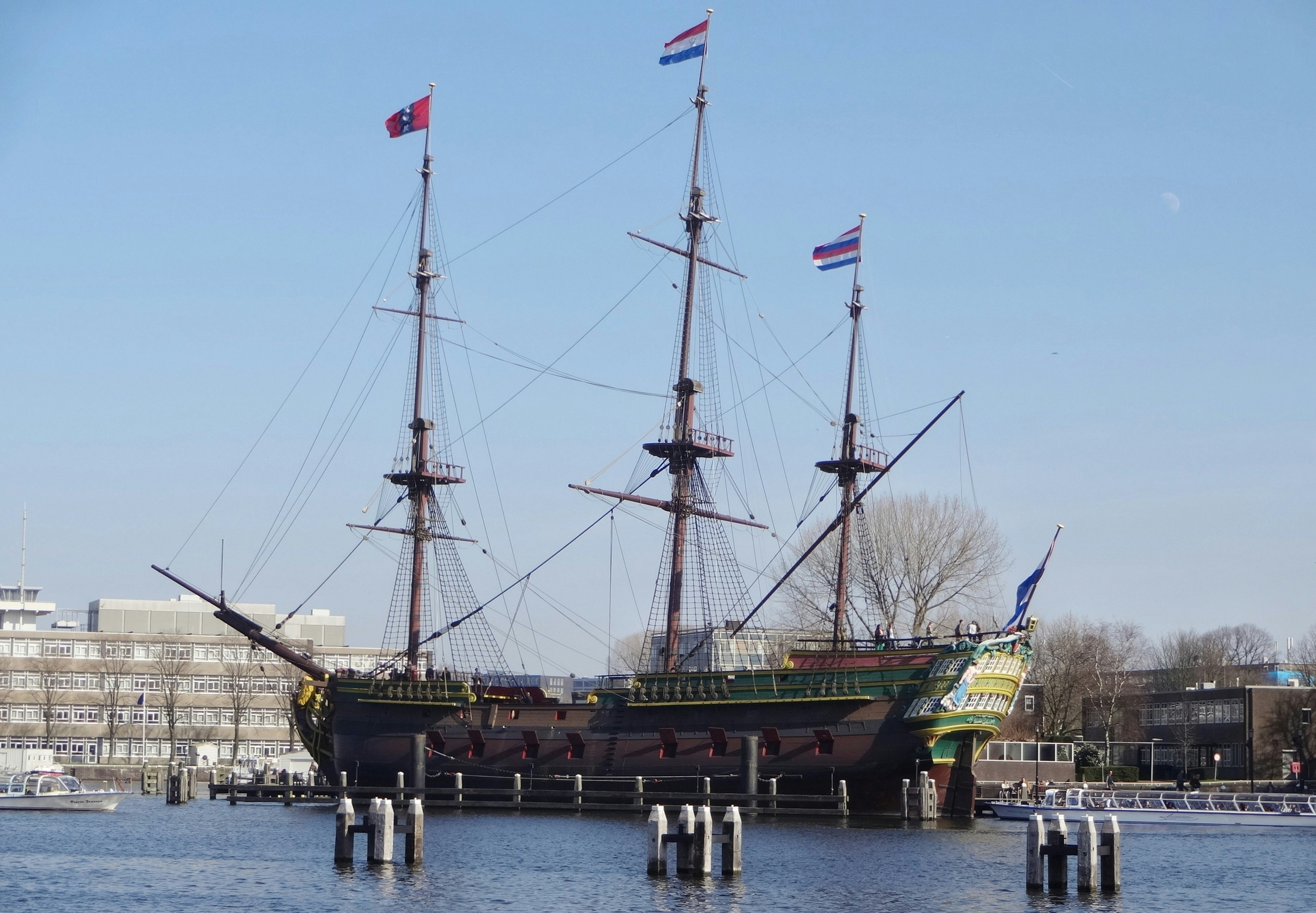 Three-masted ship docked by calm water beneath a clear blue sky, its rigging and dark hull reflected in the harbor.