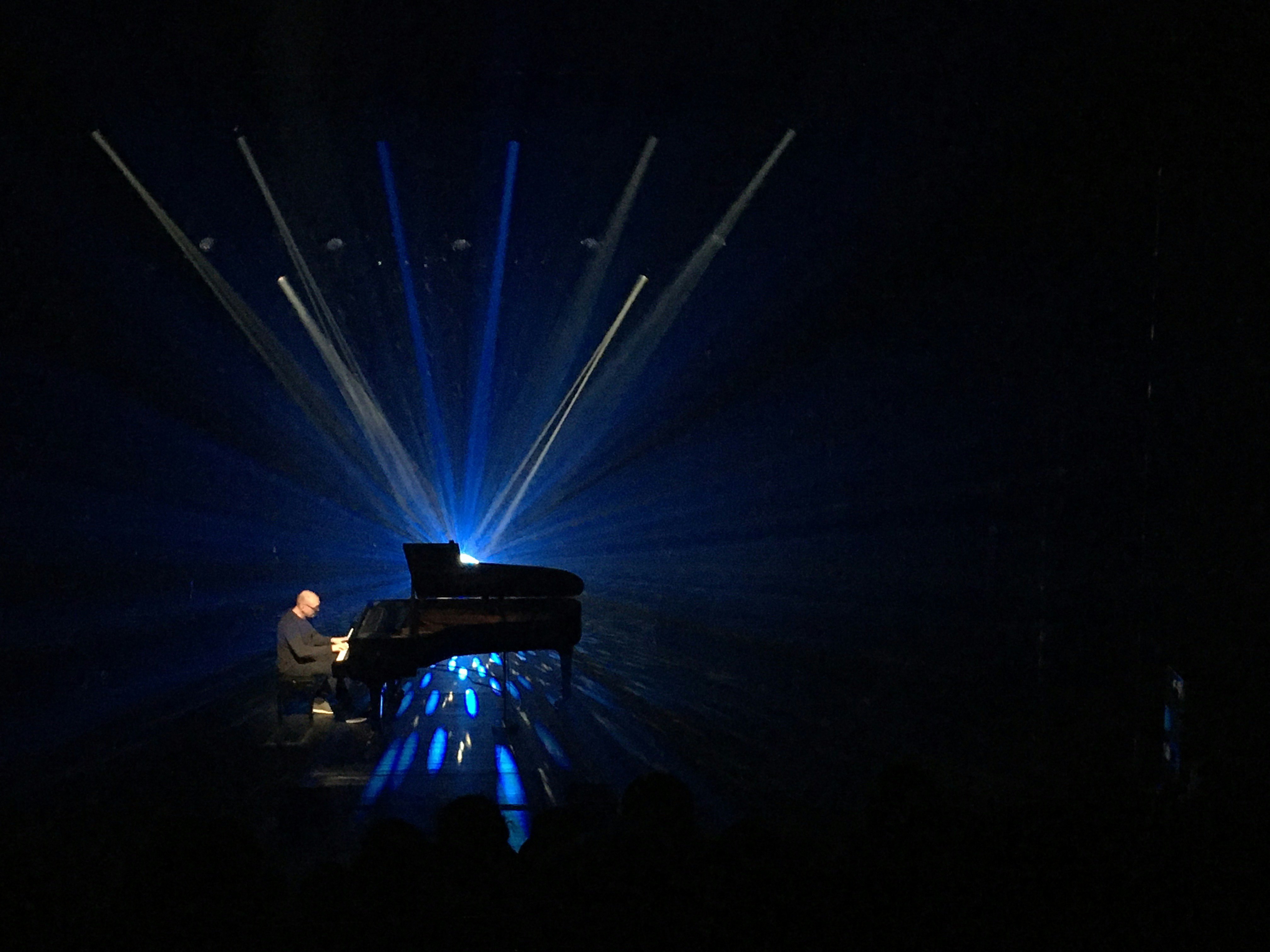 A pianist performs passionately at a grand piano, surrounded by beams of blue light cutting through the darkness.