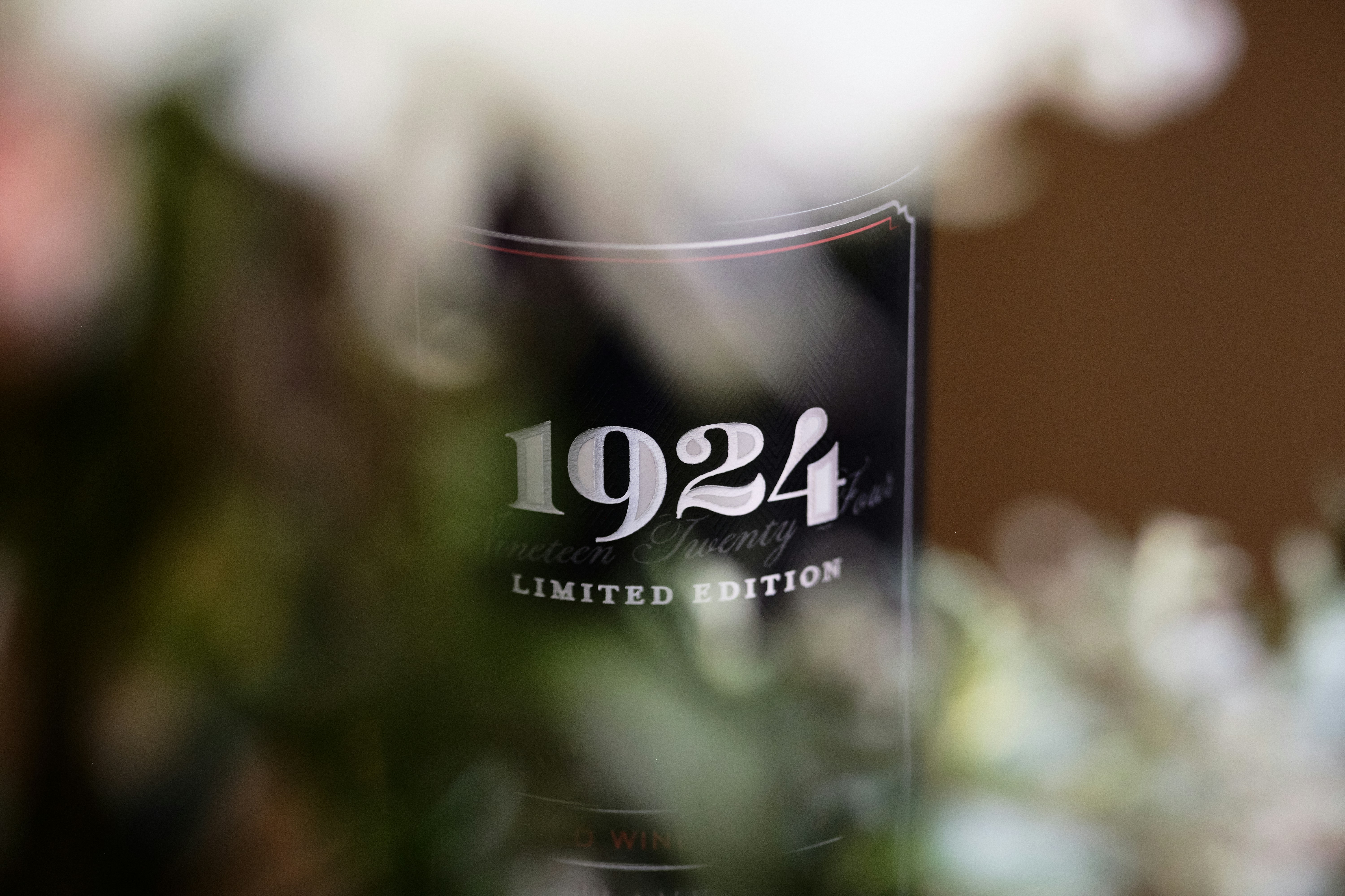 A blurred close-up of a limited edition bottle with the year '1924' visible, surrounded by soft floral bokeh. The focus is intentionally shallow to emphasize the label's glow.