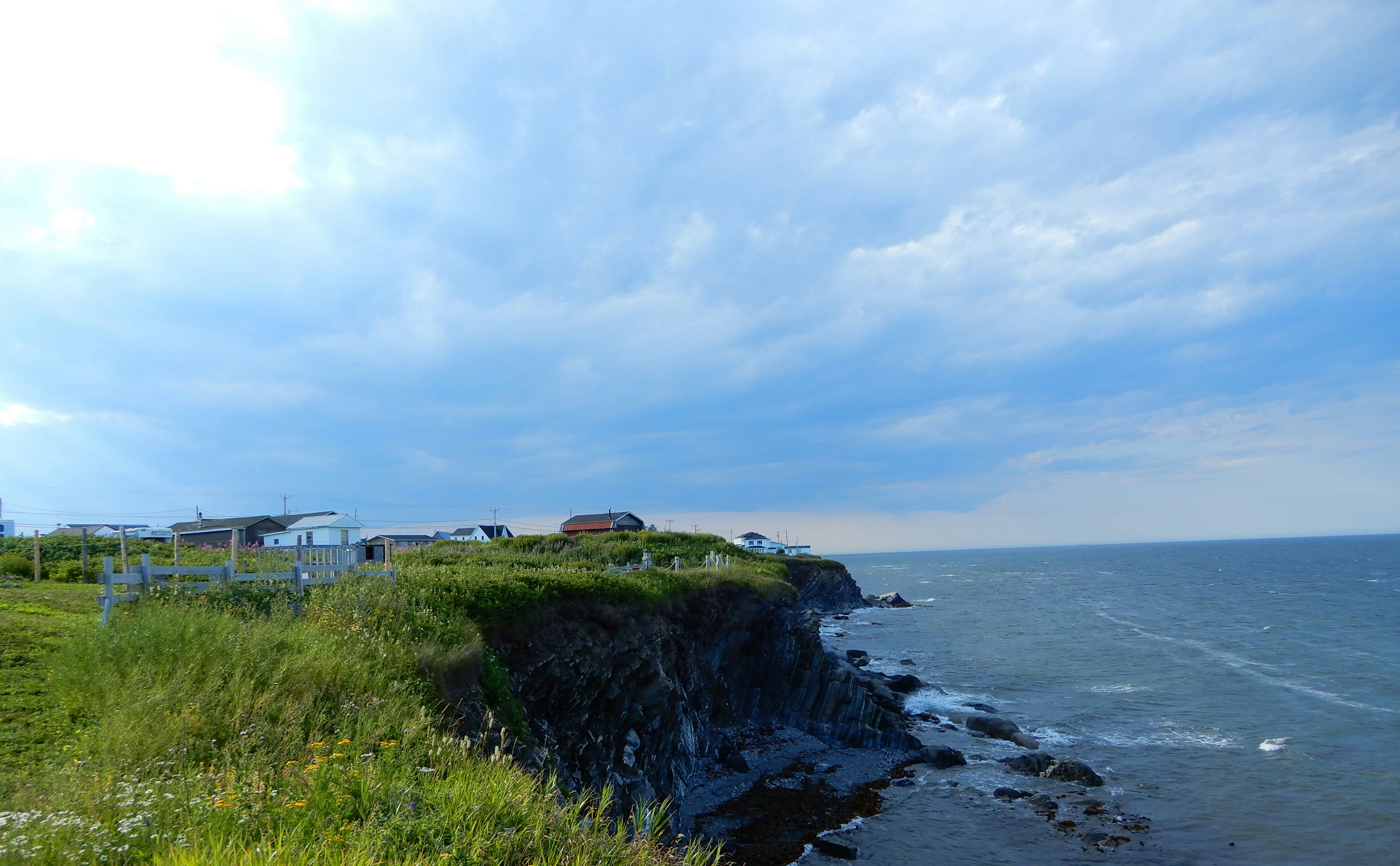 Photograph capturing clifftop houses overlooking a rocky shoreline, with grassy foreground and a calm sea under a cloudy blue sky.