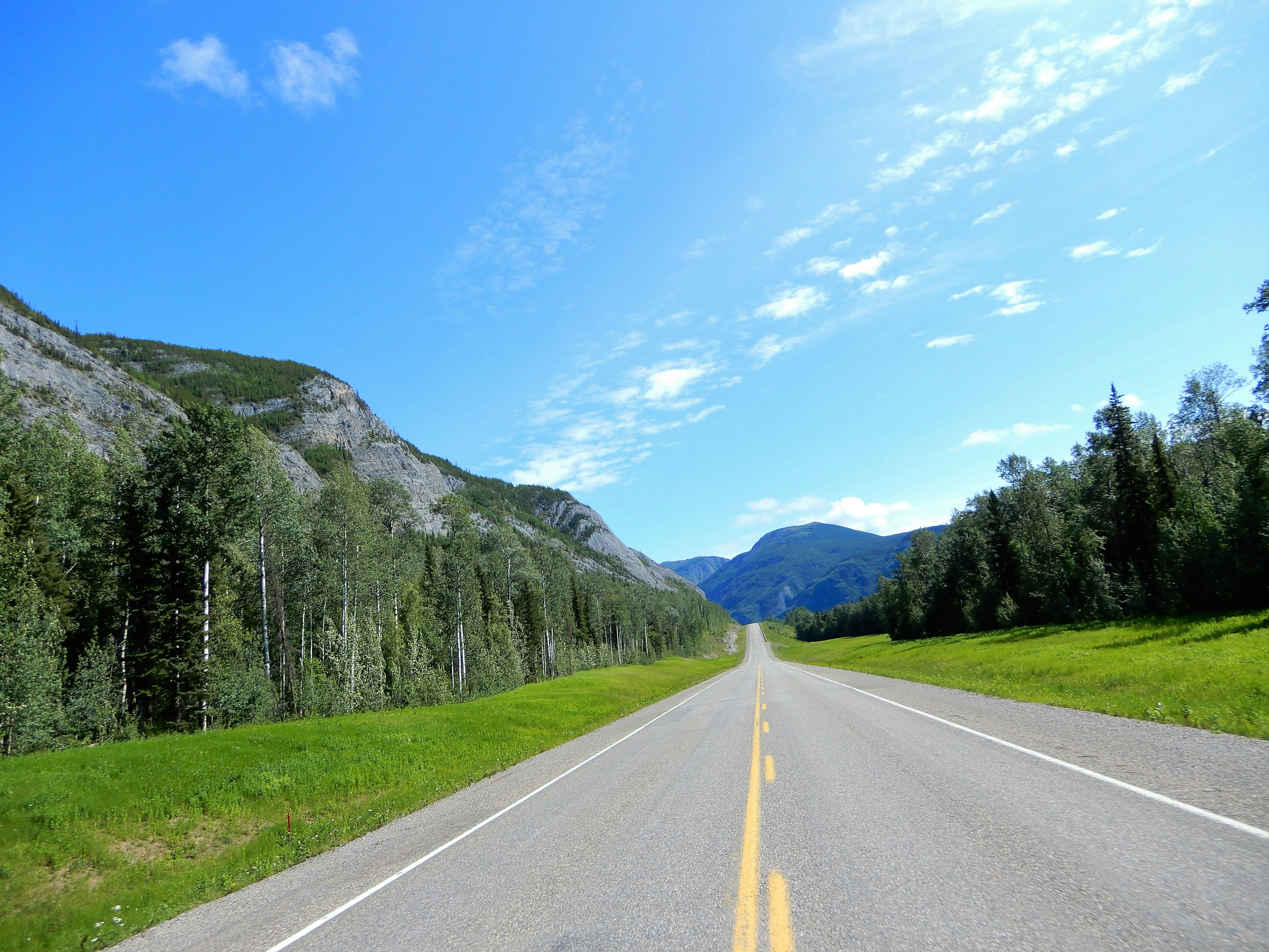 Long road flanked by lush greenery and distant mountains under a bright blue sky.