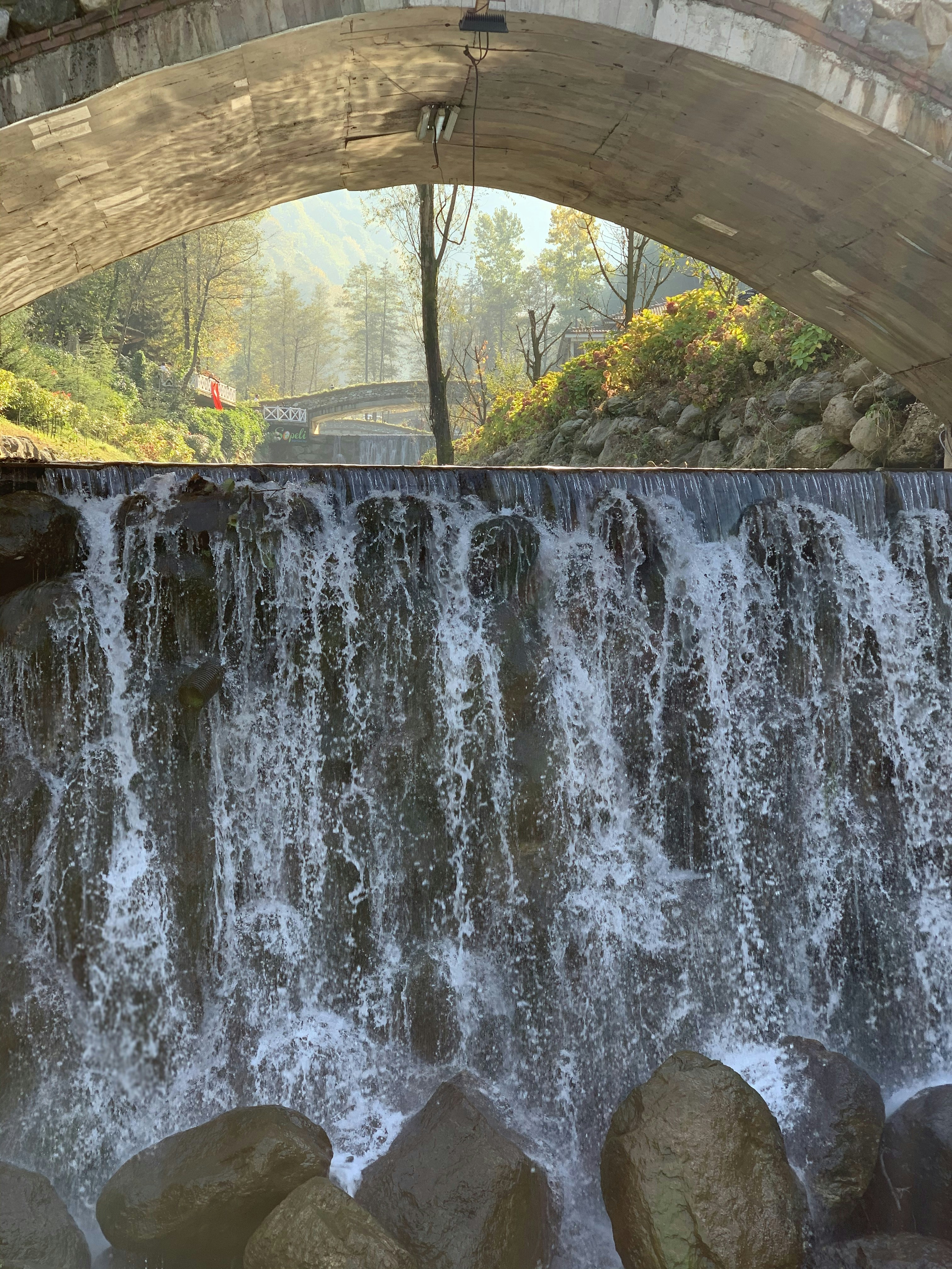 Flowing waterfall cascading over rocks beneath a stone bridge, surrounded by autumn foliage.