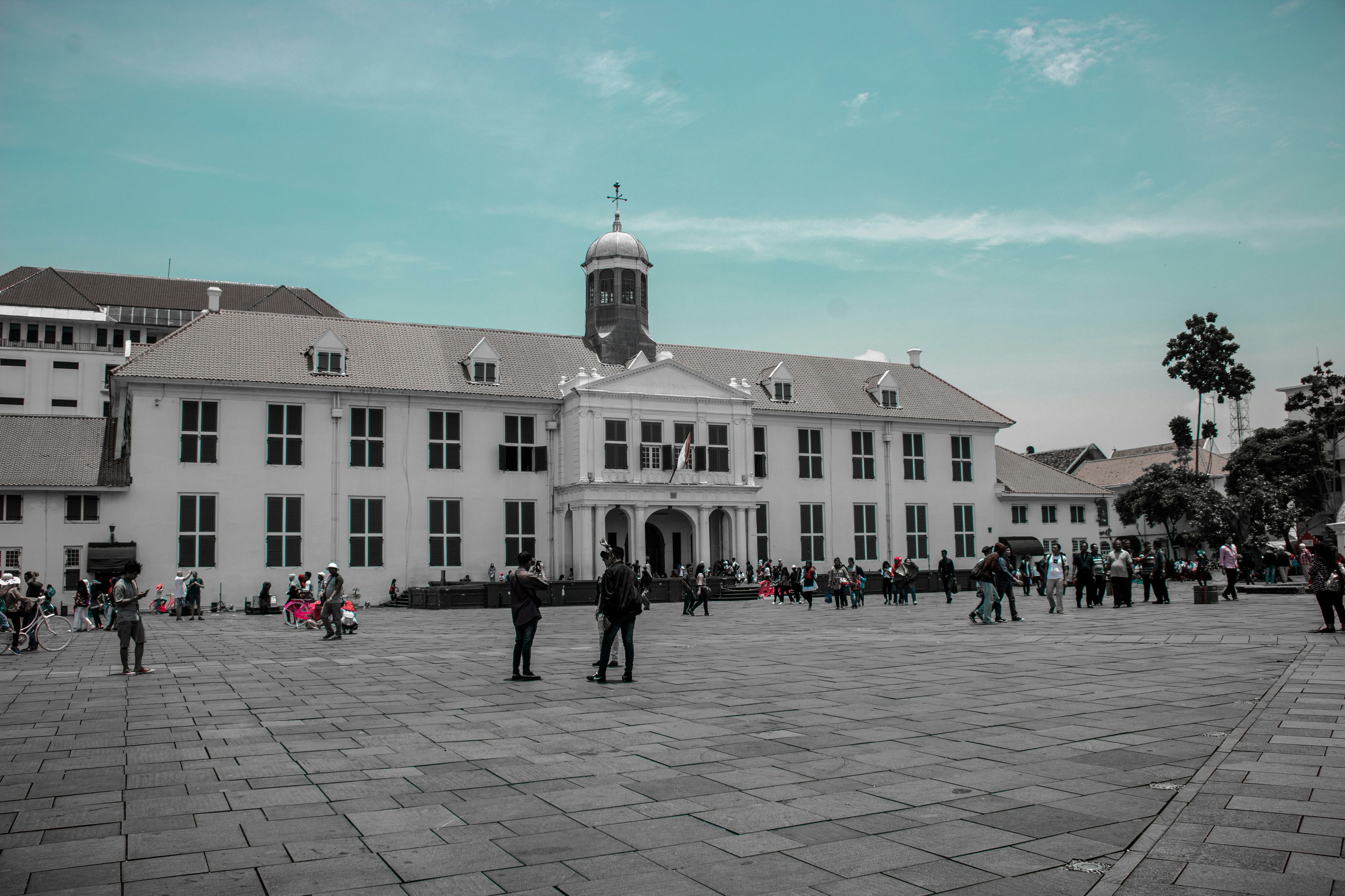 People walking across a large plaza in front of a historic building under a bright blue sky.