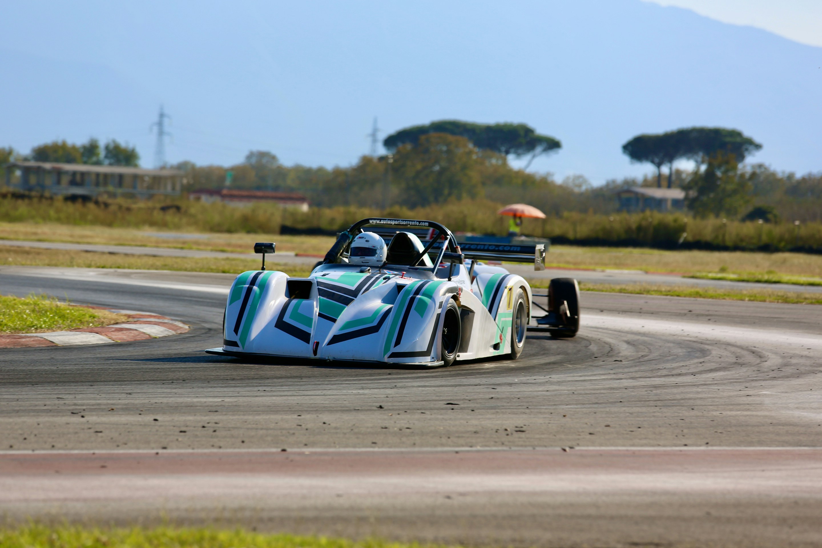 A sleek race car navigates a curve on a racetrack, showcasing dynamic motion and vibrant design elements. The background features trees and distant hills.