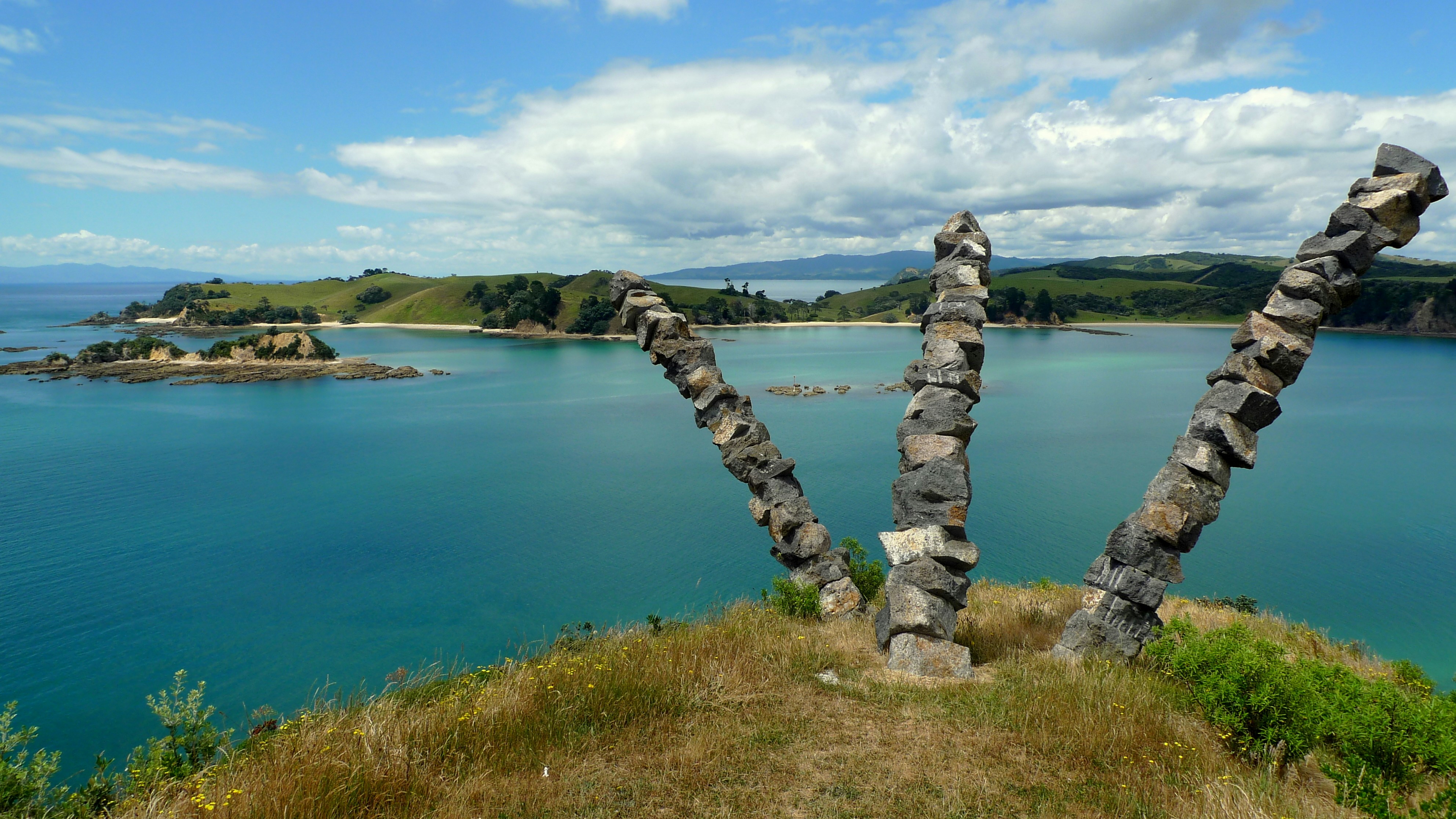 Three towering stone pillars stand against a backdrop of tranquil blue waters and lush green hills, highlighting the harmony between nature and artistic expression.