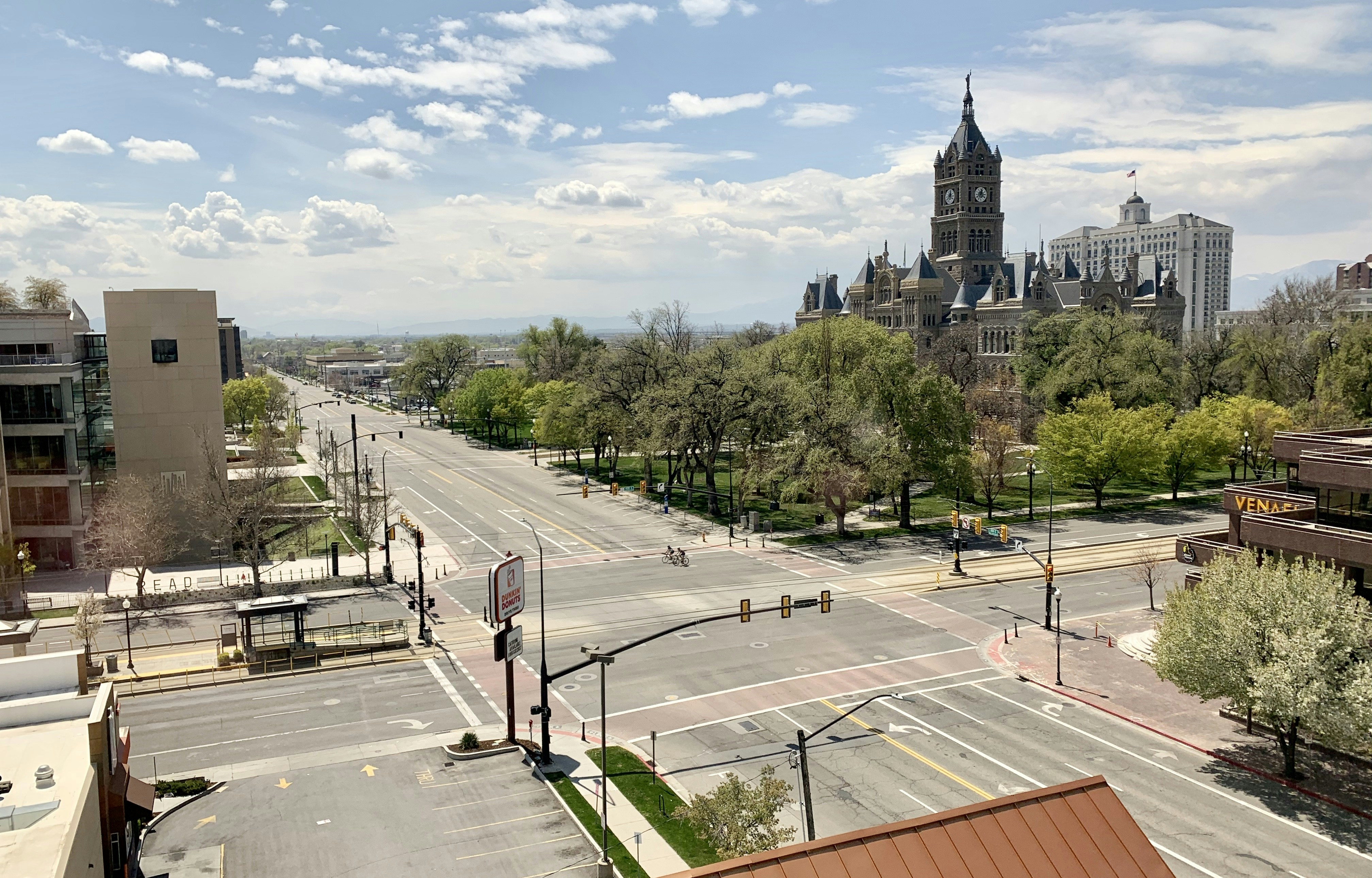 Wide city street lined with lush green trees and historic buildings under a bright blue sky.