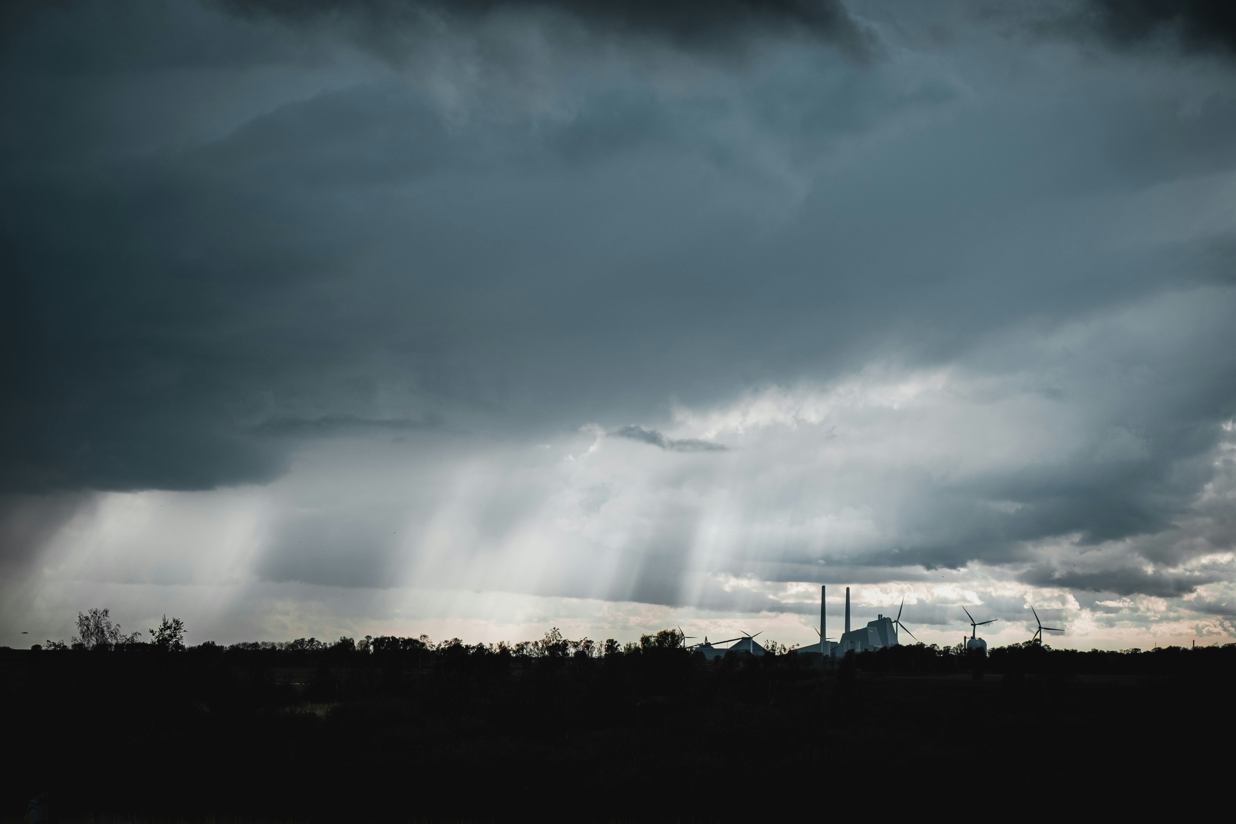 Dramatic clouds parting to reveal rays of light illuminating a distant industrial landscape. The contrast between nature and man-made structures is striking.