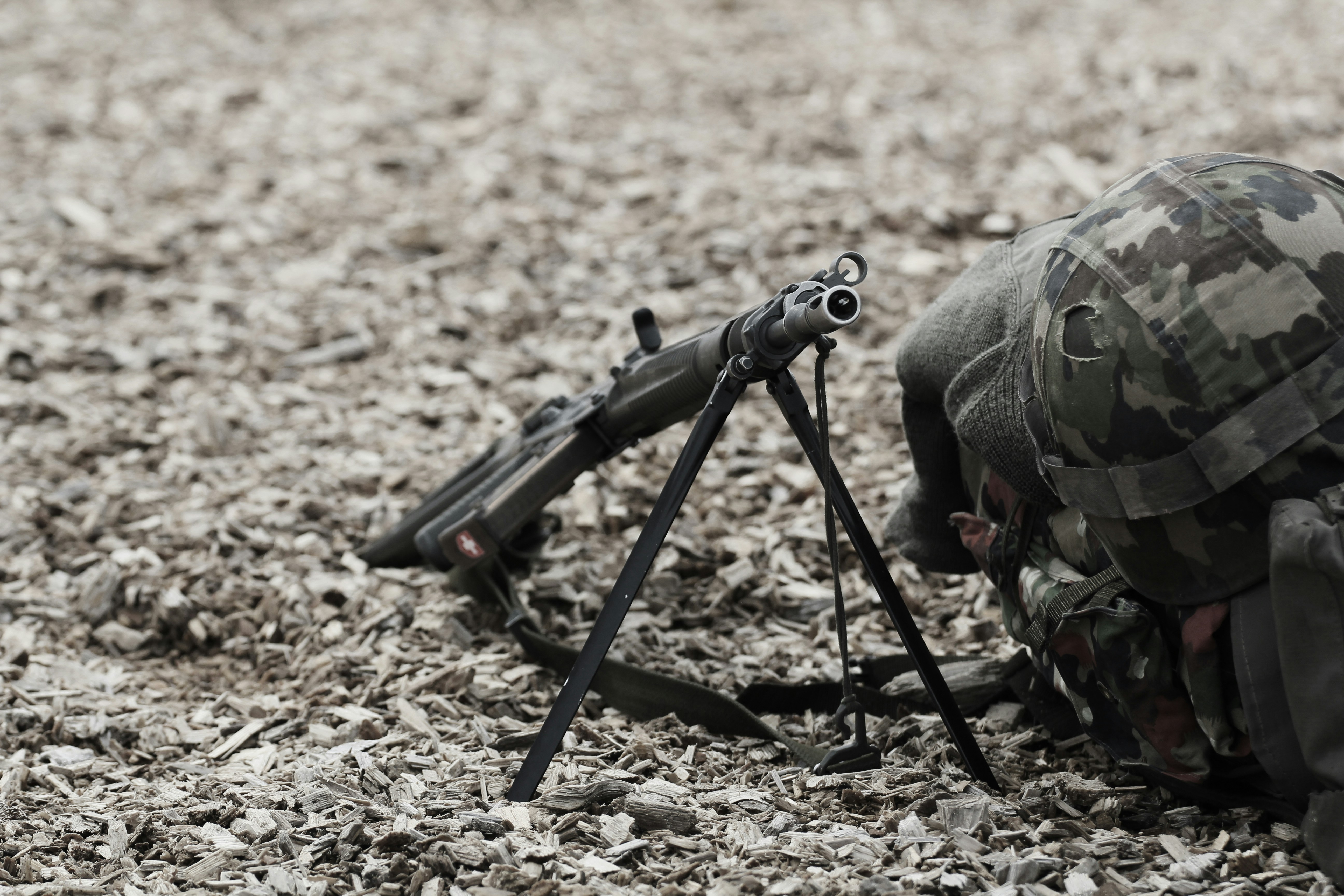 A soldier in camouflage crouches beside a rifle set on a bipod, surrounded by a textured landscape of wood chips. The scene conveys a sense of focus and readiness.