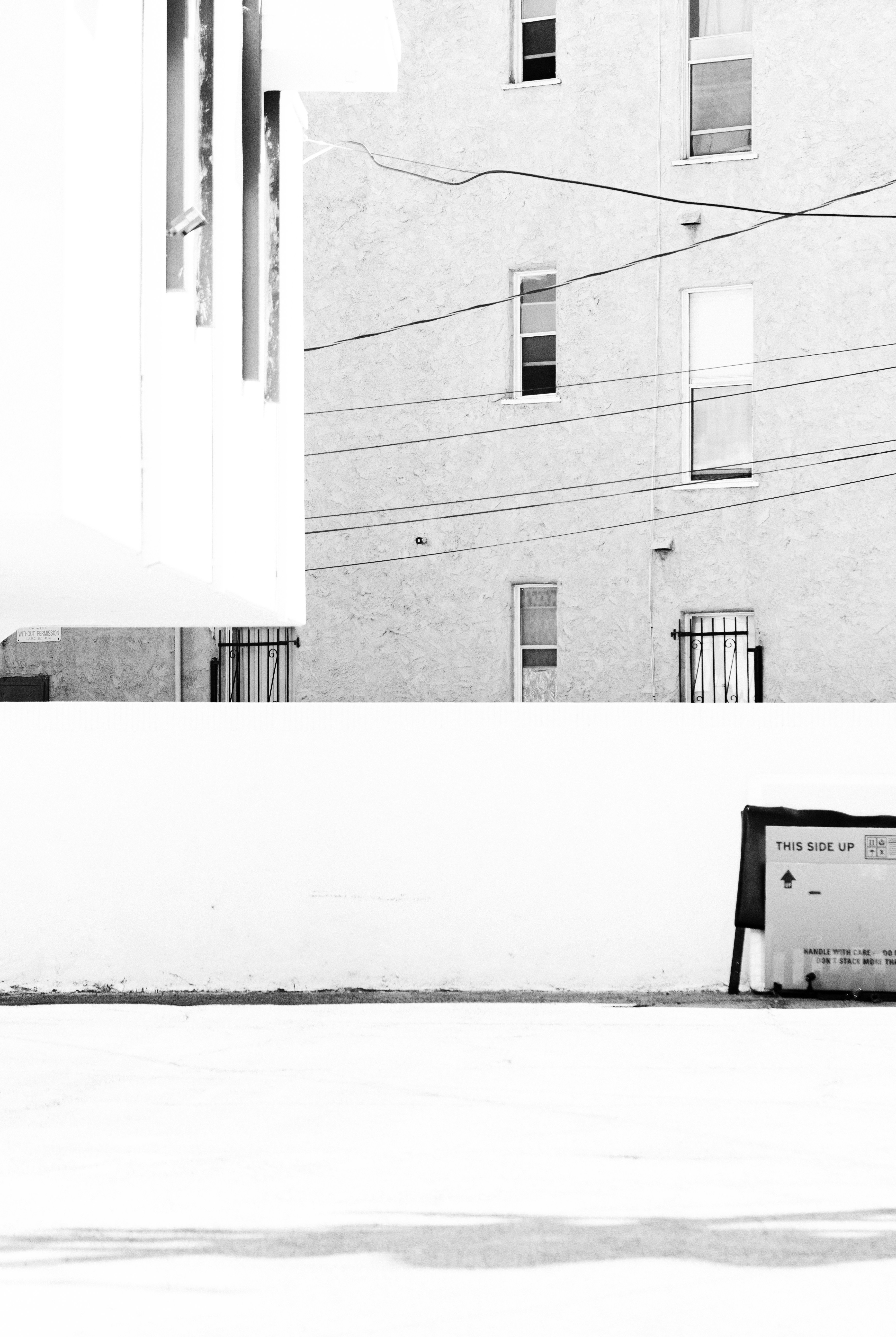 A monochromatic scene featuring a stark white wall and a cardboard box marked 'THIS SIDE UP,' set against a backdrop of a textured building with windows and power lines.
