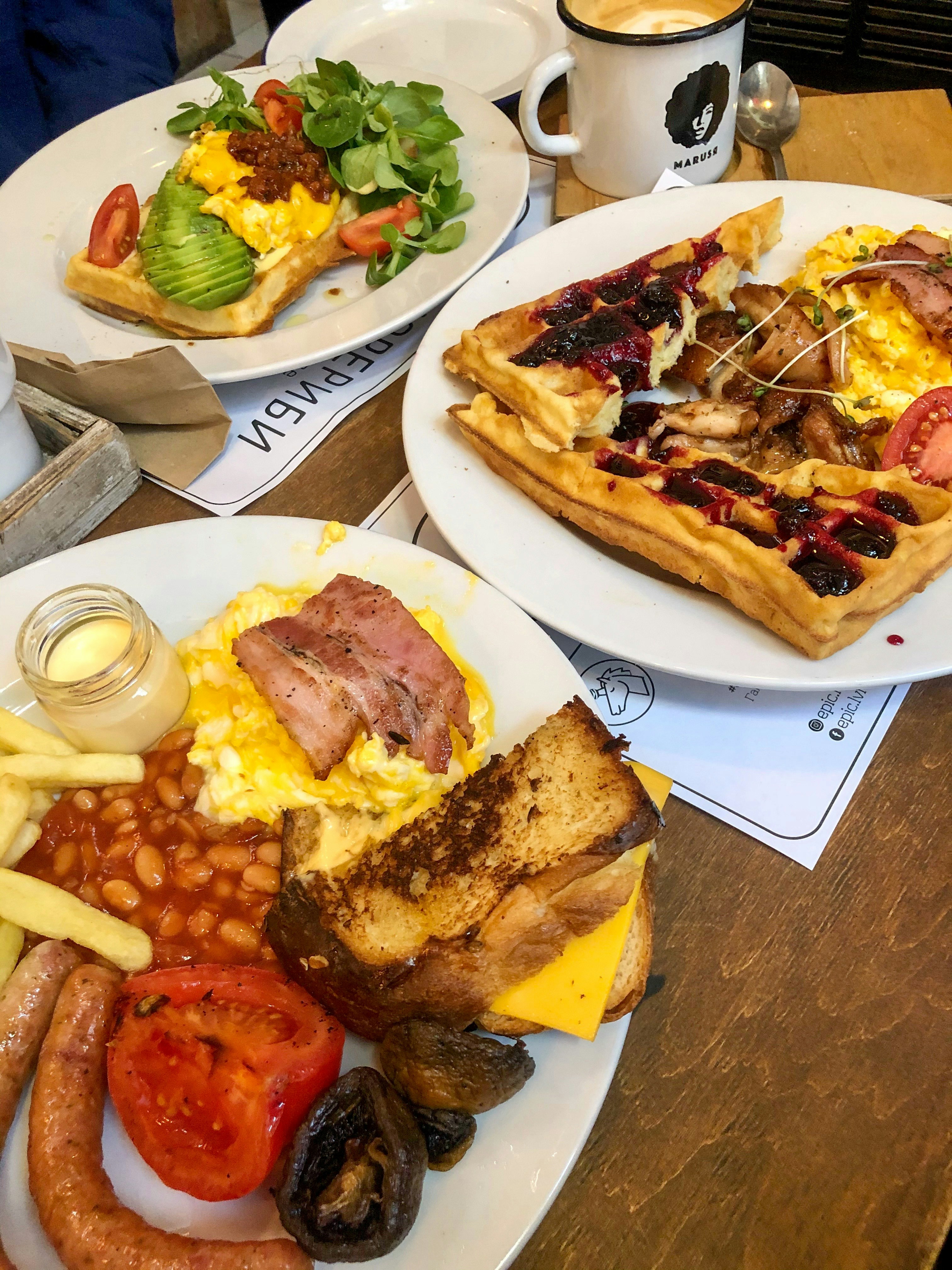 Three-Plate Brunch Mosaic: Avocado toast, berry-topped waffle, and a hearty egg-and-sausage plate on a rustic cafe table