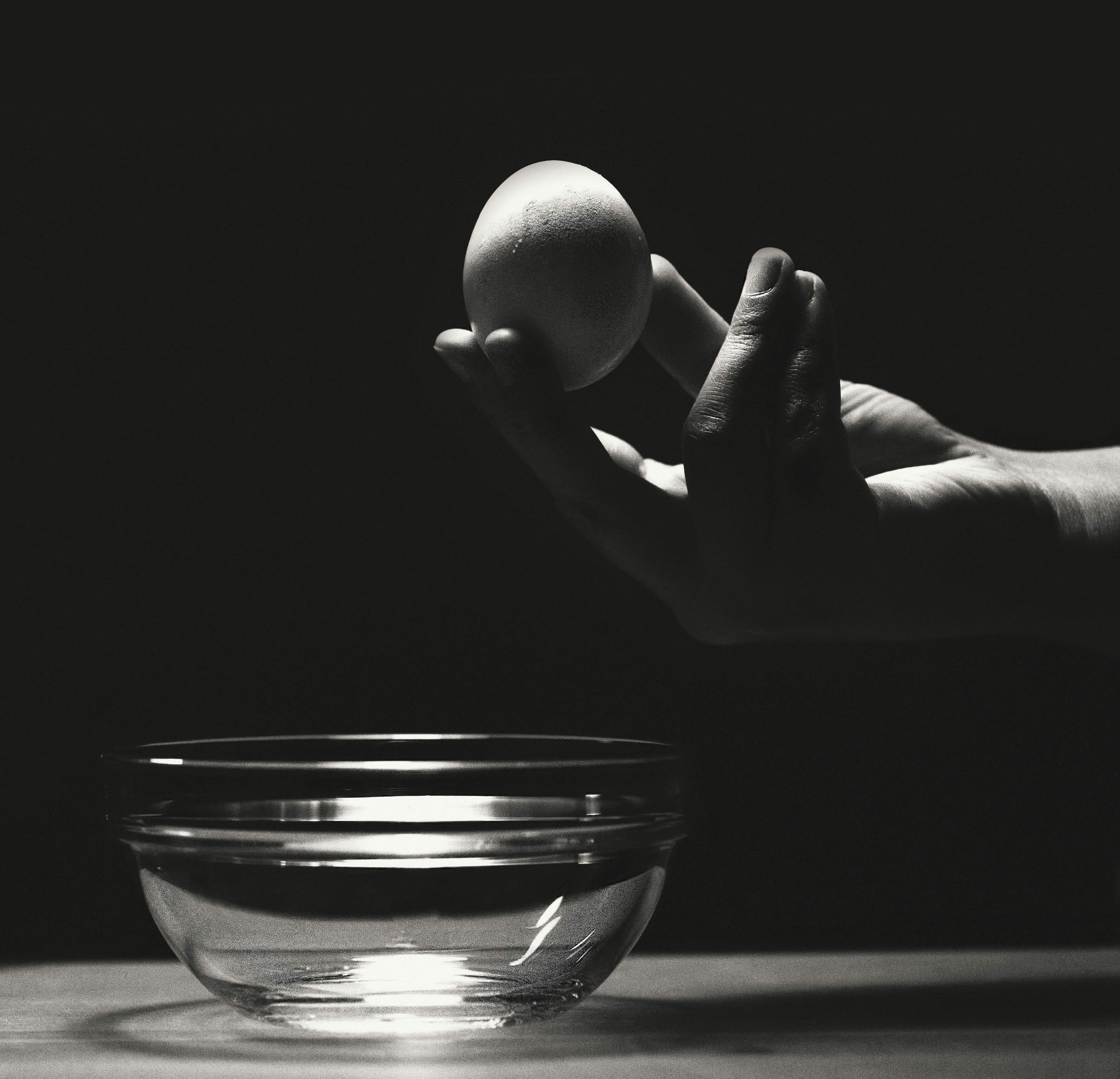 A hand poised delicately above a glass bowl, holding a single egg, set against a stark black background.