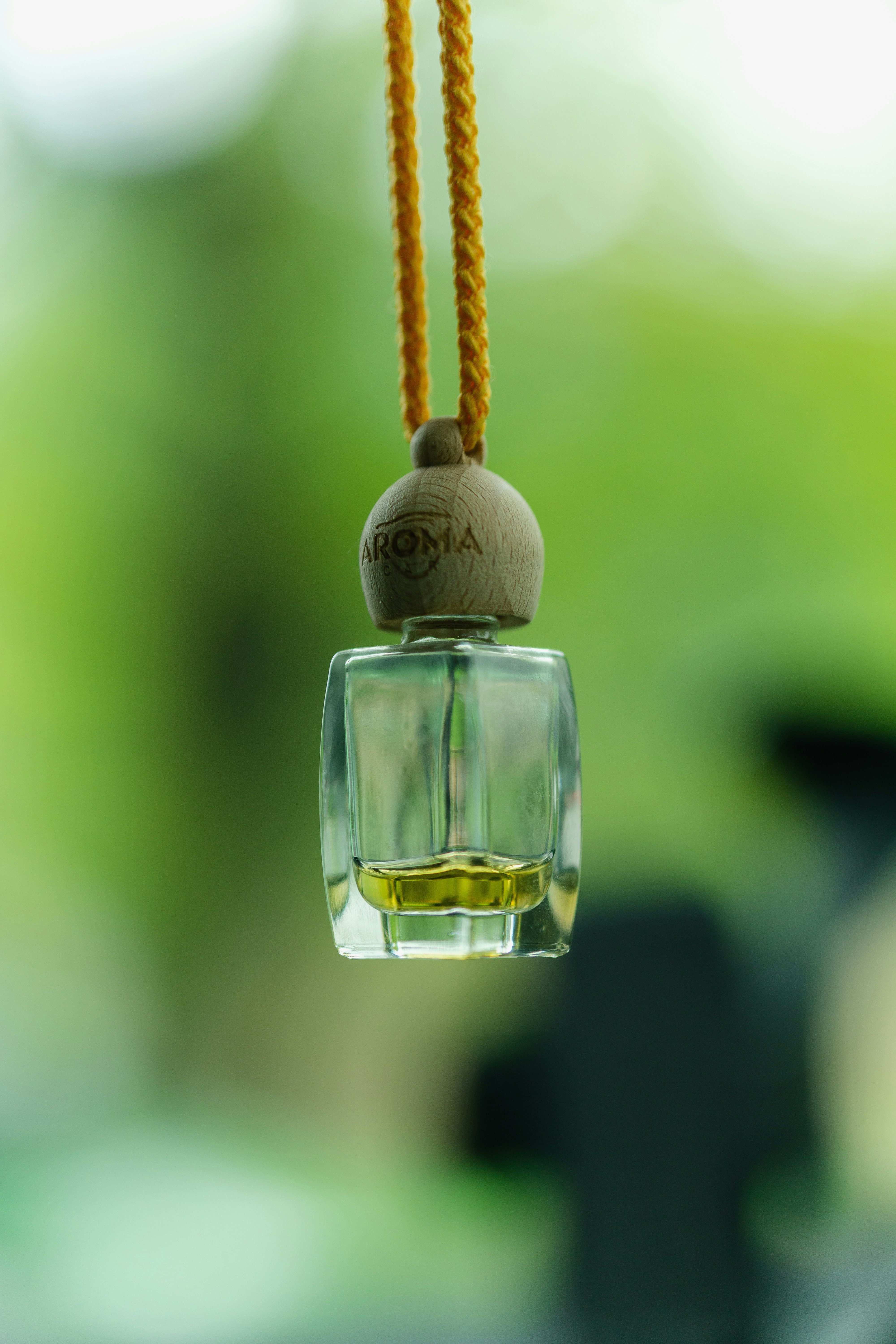 Aromatic pendant hanging against a softly blurred green background, showcasing its glass container filled with a golden liquid. The wooden top features an engraved logo.