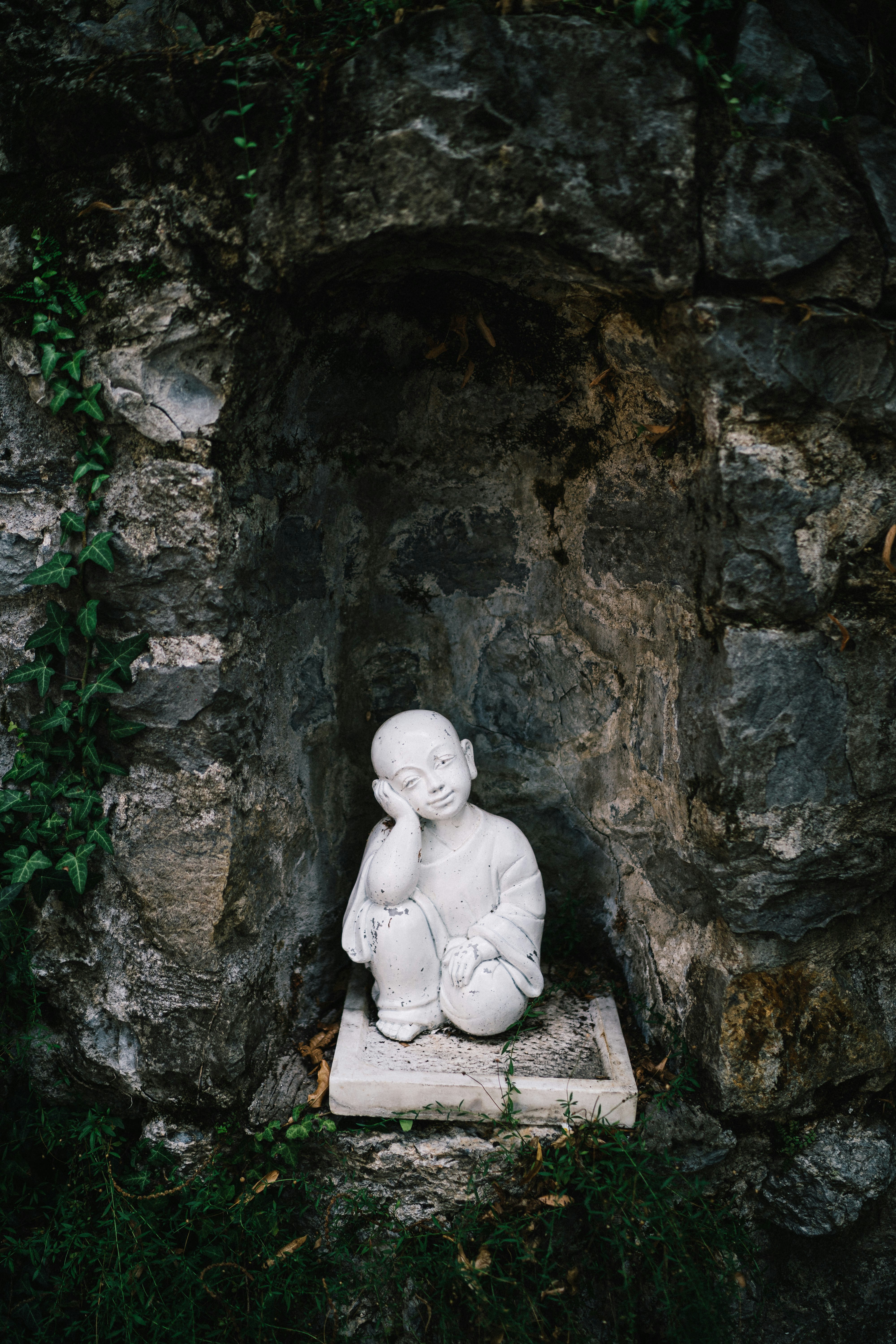A serene white statue of a child monk resting against a stone wall, surrounded by creeping vines and natural textures.