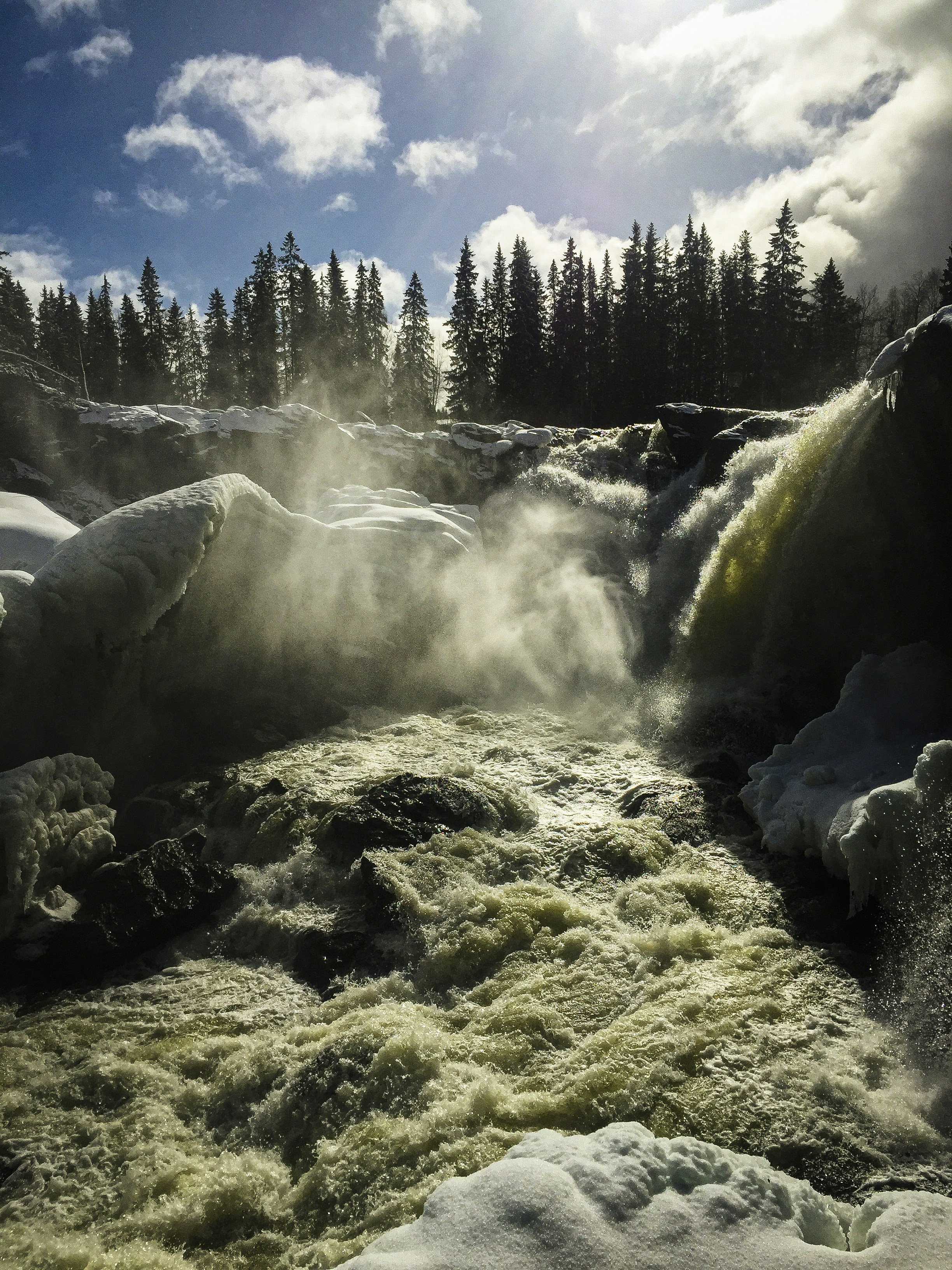 Waterfall cascading between snow-covered rocks with mist rising into a sky framed by evergreen trees.