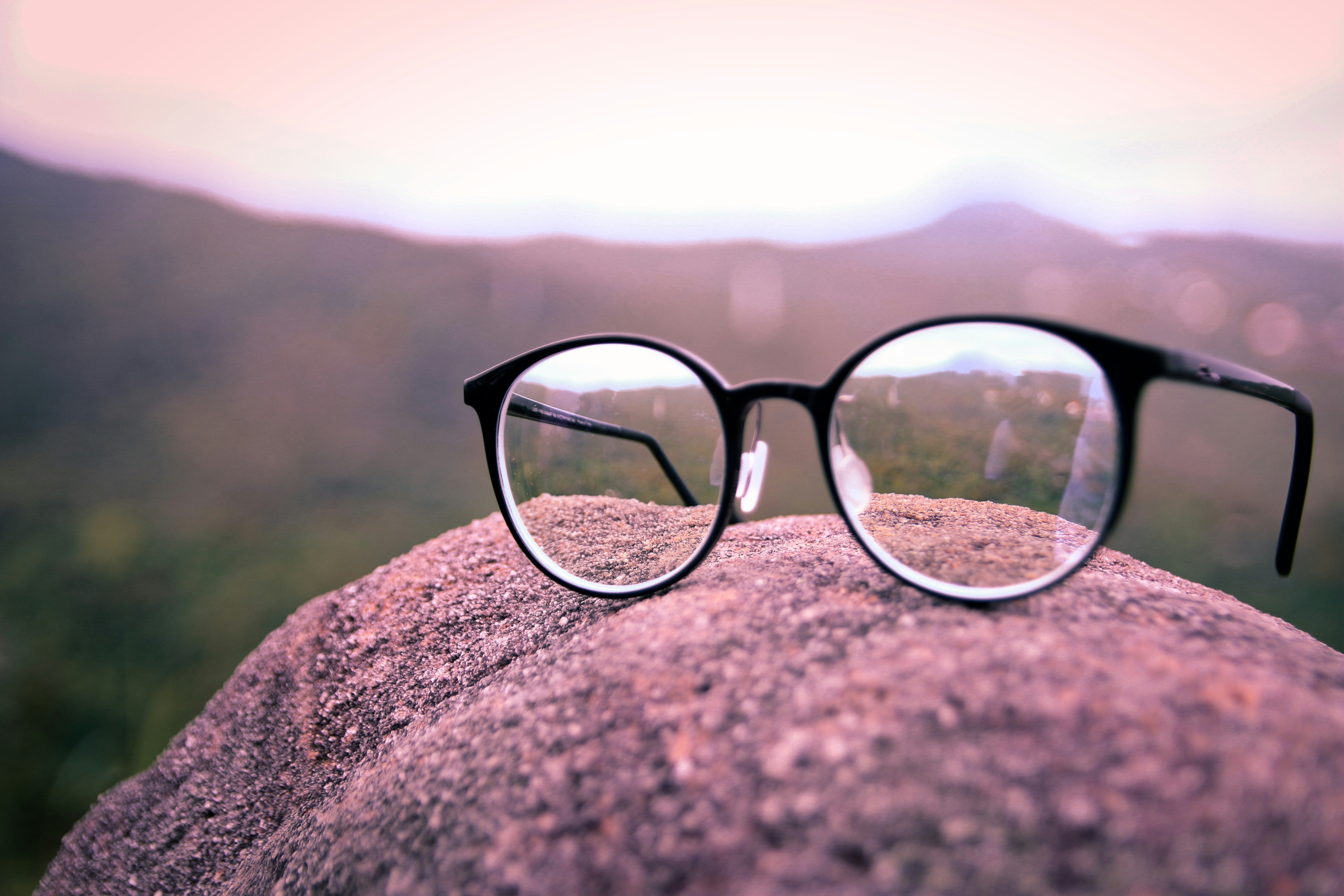 A pair of glasses resting on a rock, reflecting a vibrant landscape in the lenses. The scene captures a moment of contemplation amidst nature.