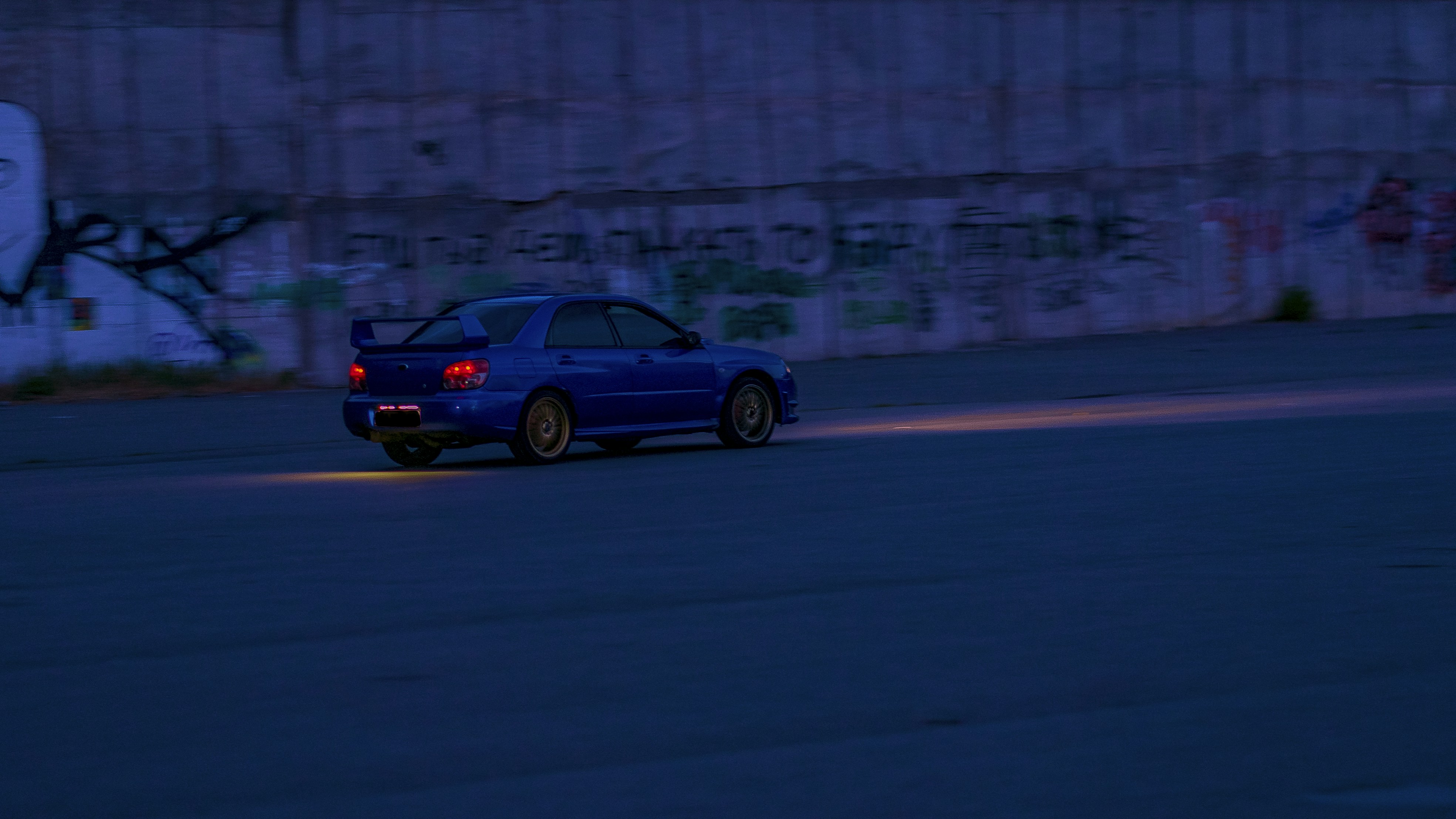 A blue sports car glides through an urban landscape, illuminated by its headlights against a backdrop of graffiti-covered walls.