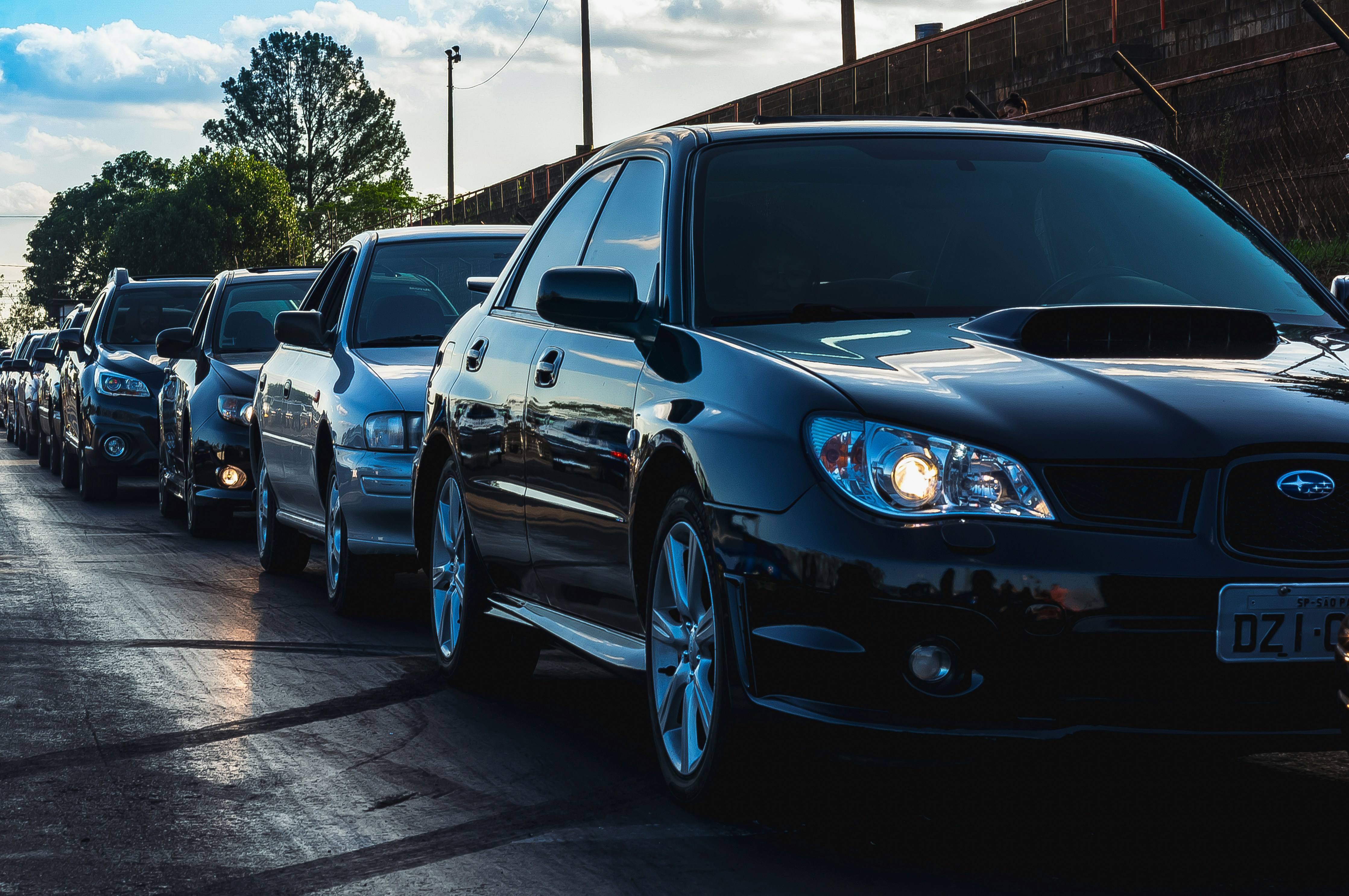 A sleek line of parked cars, featuring a prominent black Subaru in the foreground, showcasing modern automotive aesthetics against a backdrop of blue skies and distant trees.