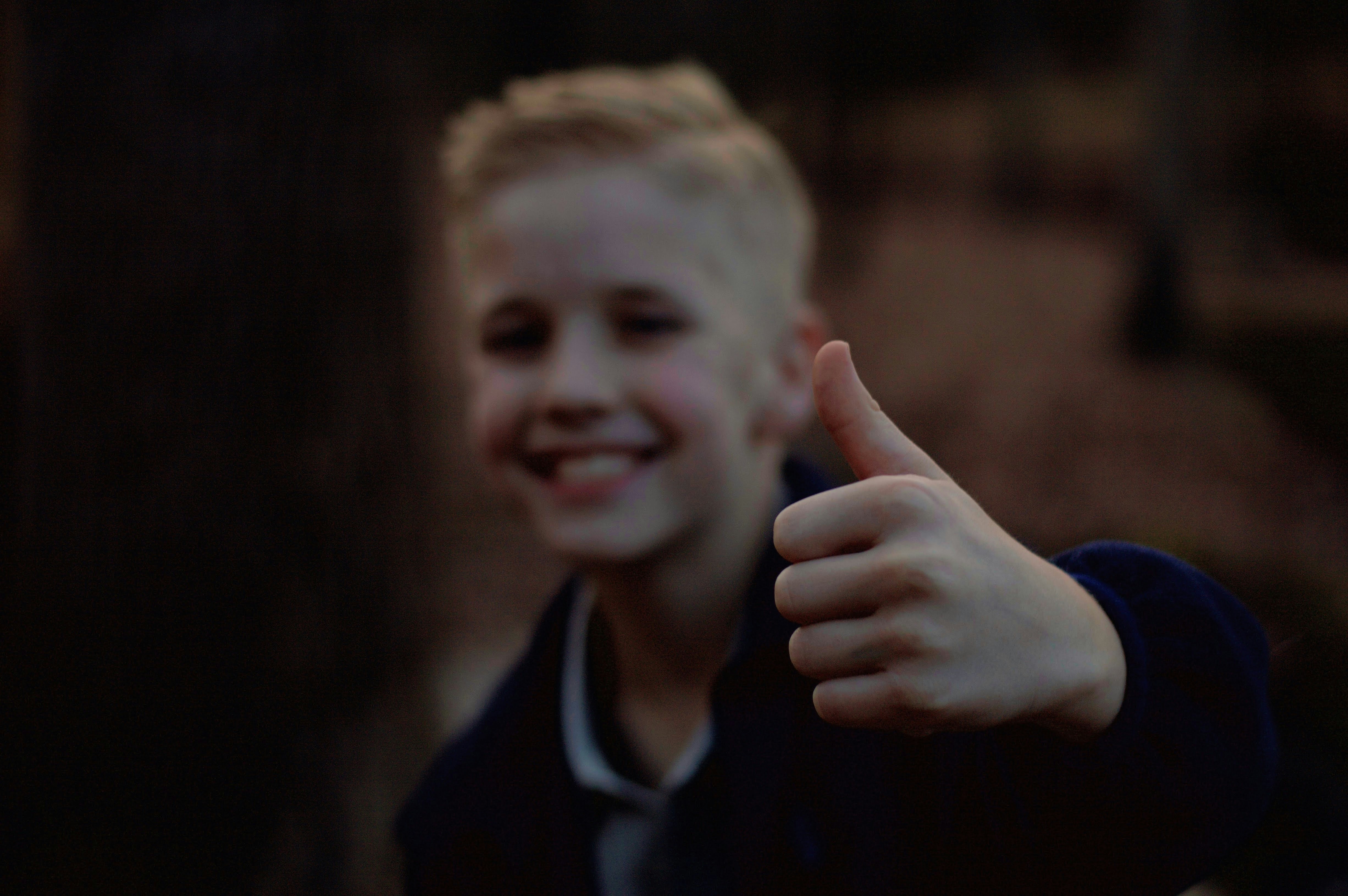 Smiling boy giving a thumbs-up gesture in low-light outdoor setting.