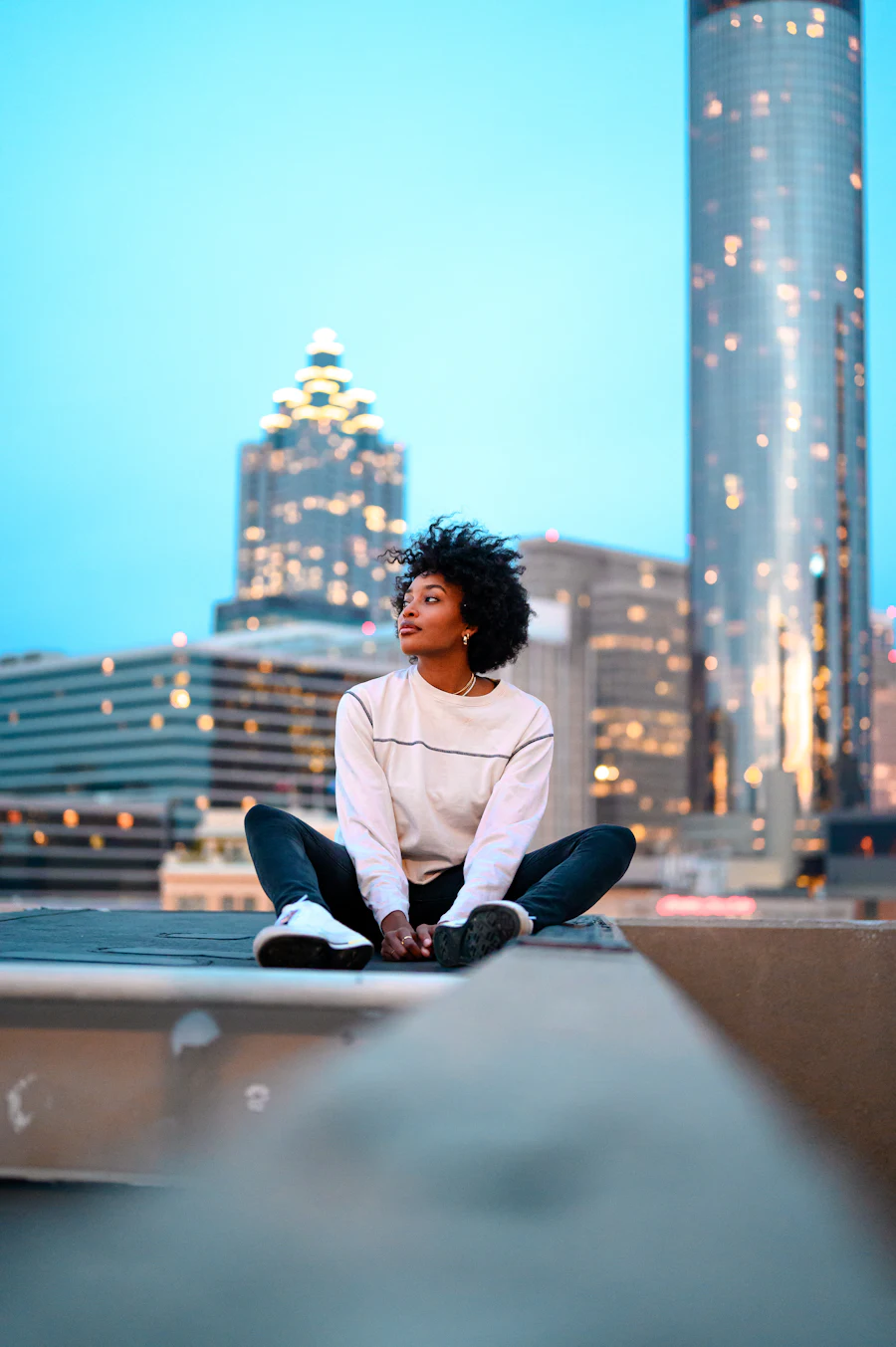 Woman sitting cross-legged on a rooftop ledge with a blurred city skyline and lit towers behind her. Soft evening light and a cool blue sky frame the portrait.