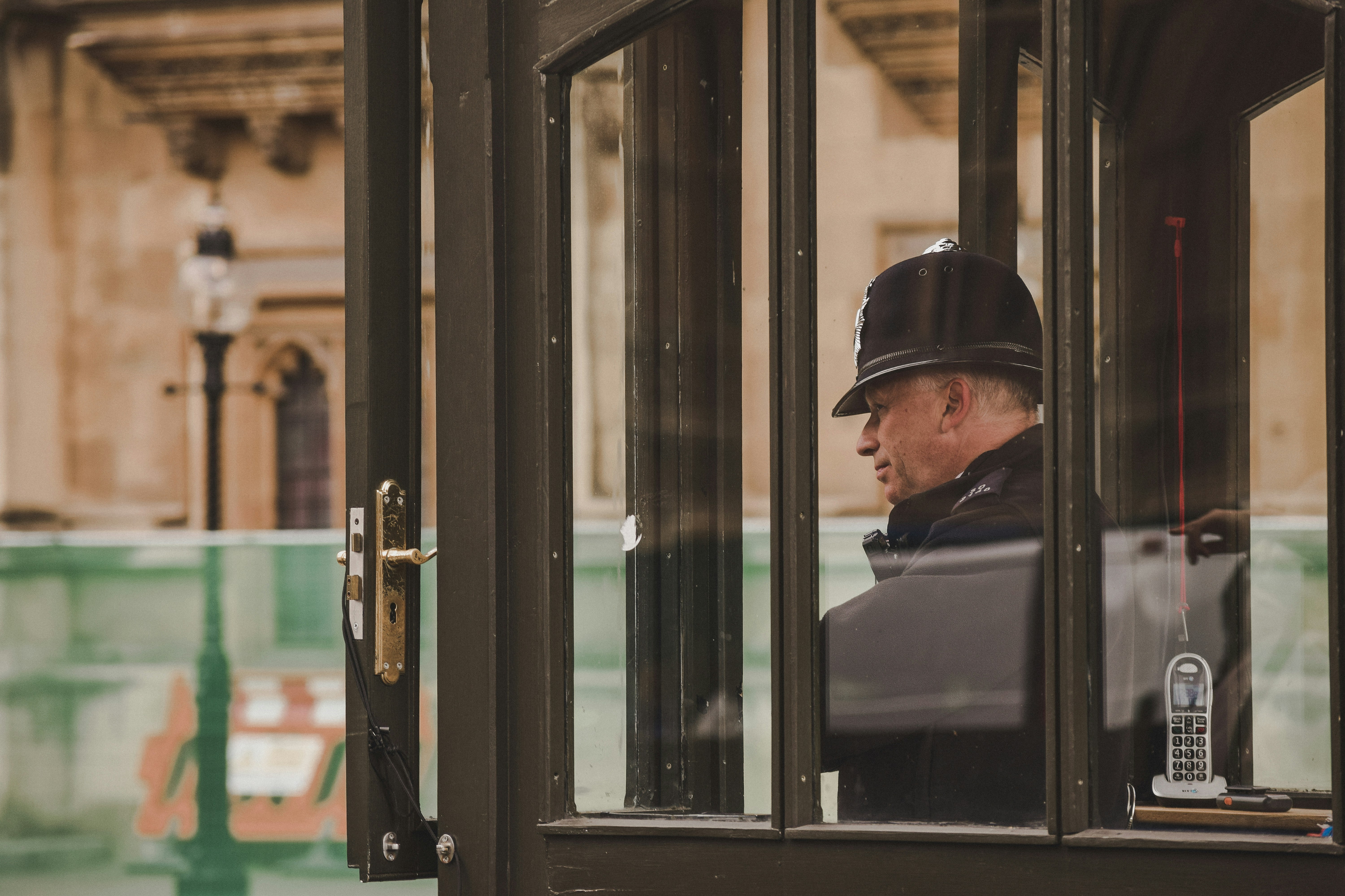 A police officer in a traditional helmet sits inside a booth, gazing thoughtfully outside. The background features blurred architectural elements, suggesting a historic setting.