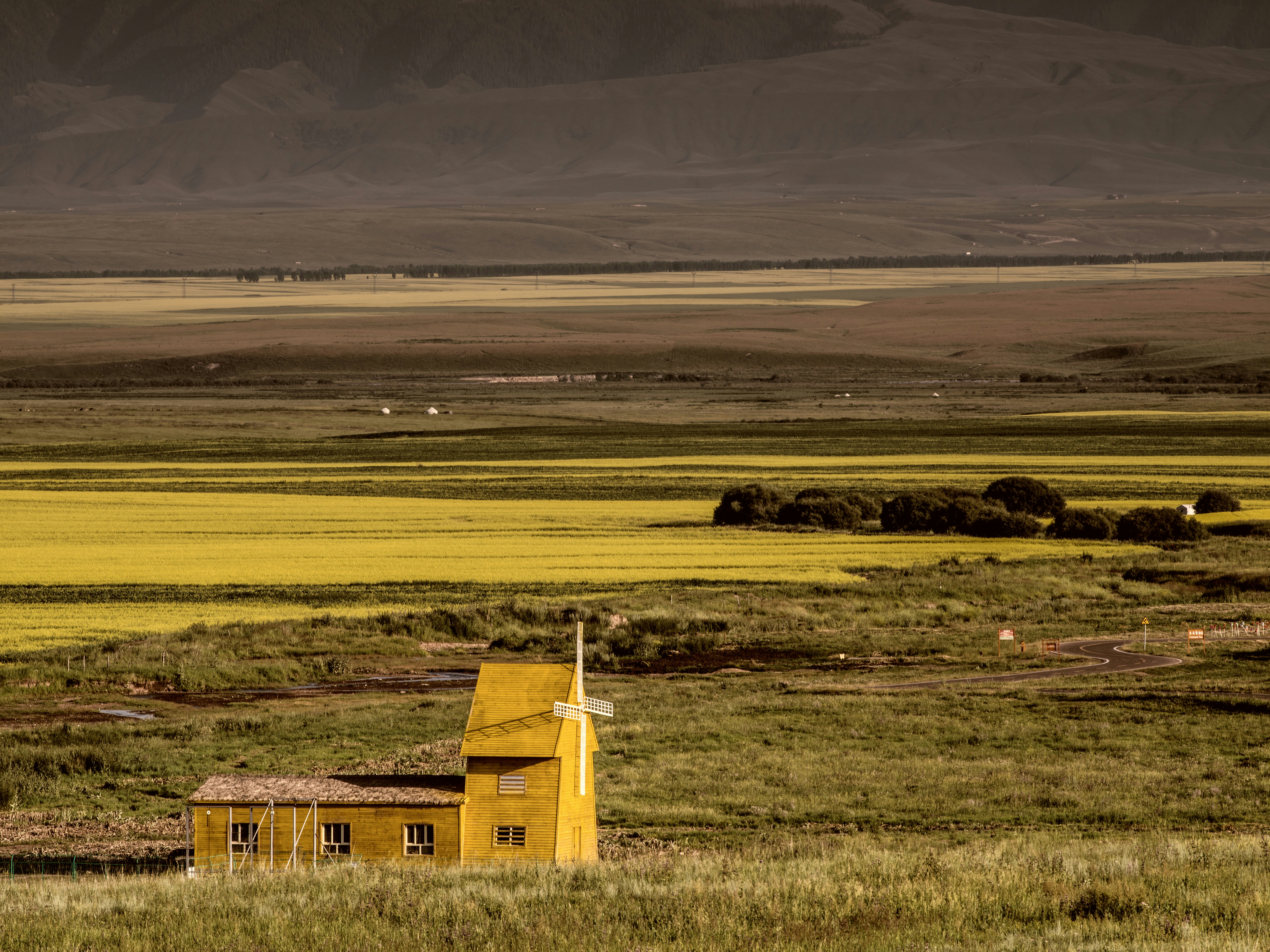 A bright yellow windmill structure stands alone in a vast green landscape, contrasting with the expansive fields of yellow flowers in the background.