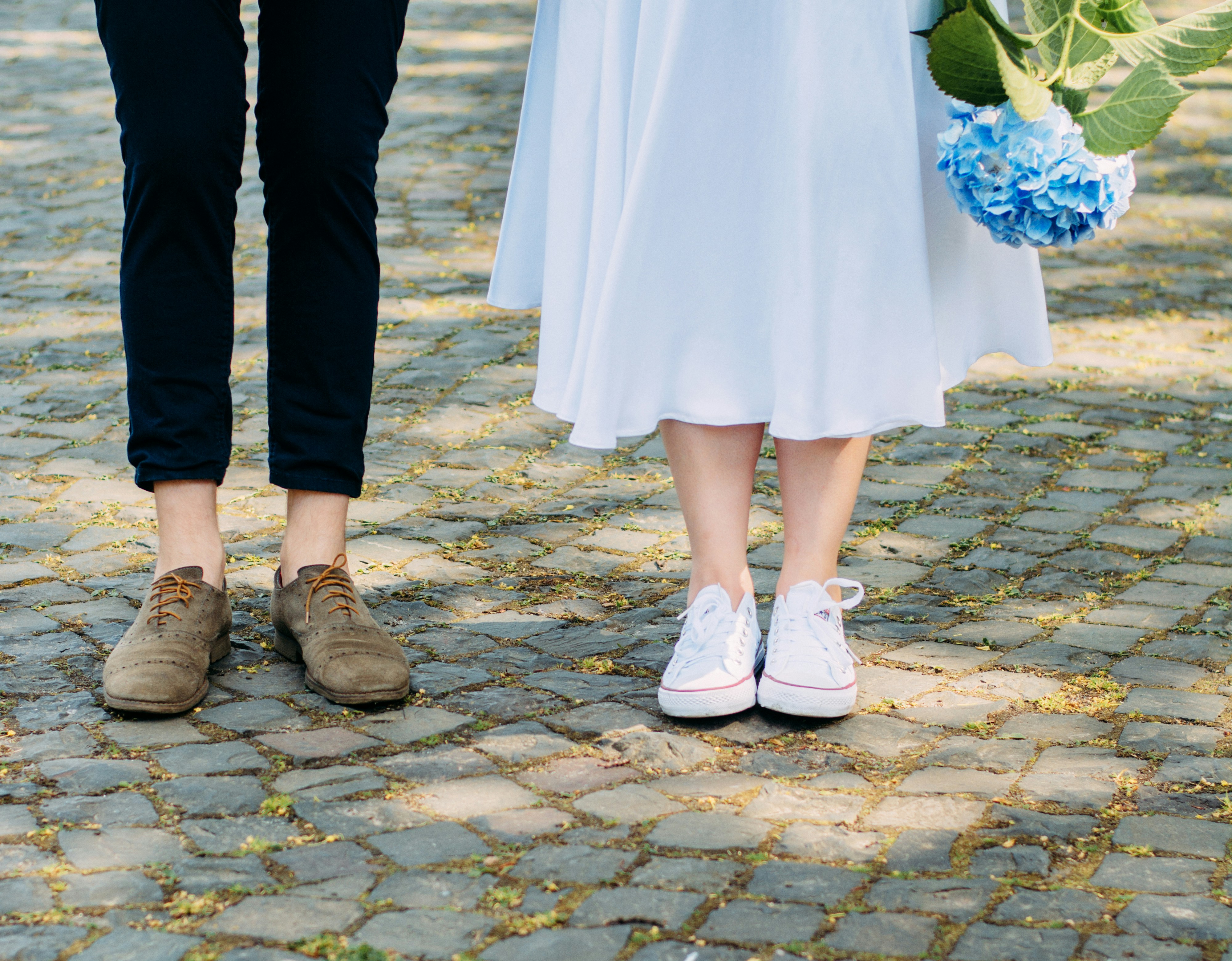 Couple's feet standing on cobblestone path, adorned with a bouquet of blue hydrangeas. The contrast of shoes adds a playful touch.
