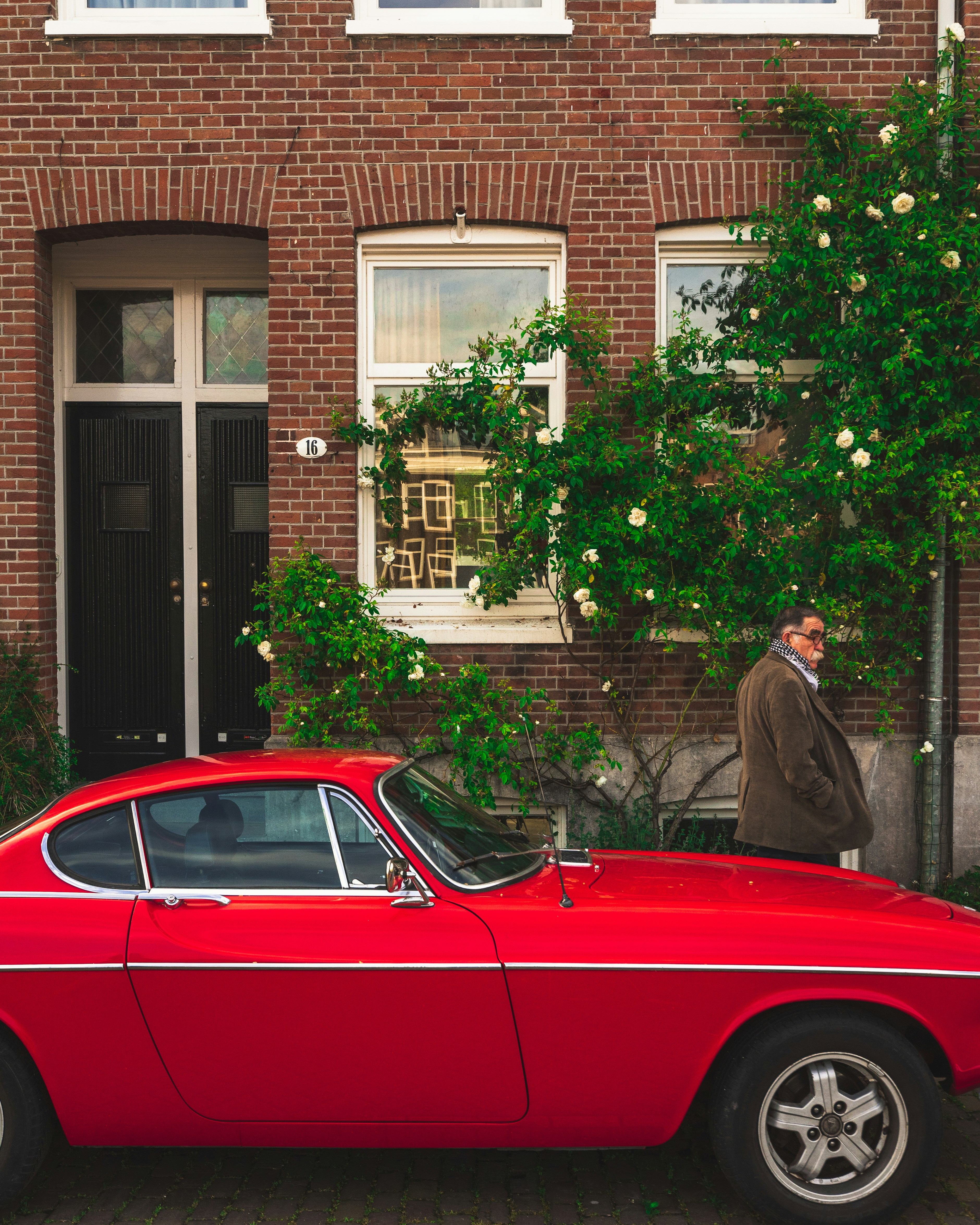 A classic red car parked beside a brick building, with a man in a scarf standing nearby, surrounded by lush greenery.