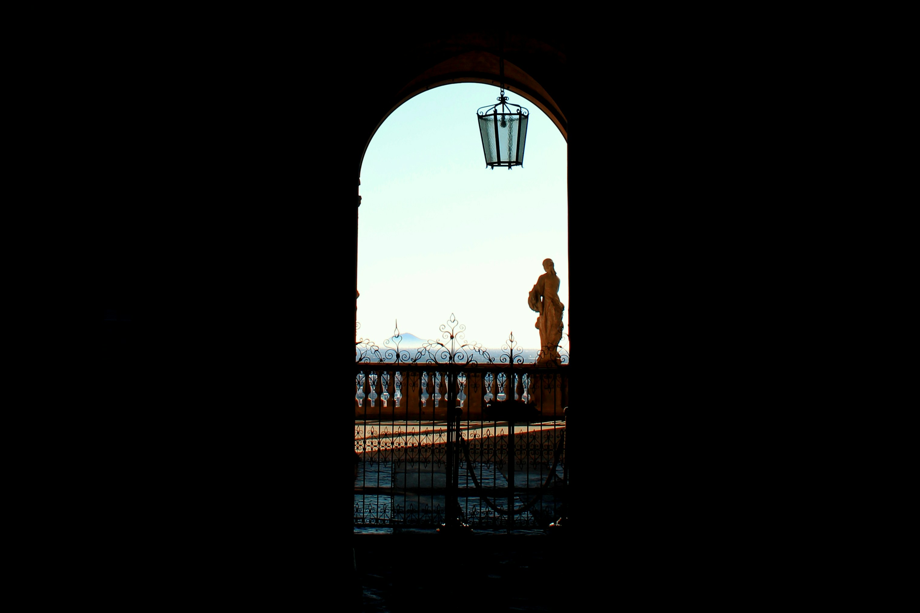 Silhouette of a statue framed by an arched doorway, revealing a serene outdoor scene beyond. The contrast emphasizes the figure's watchful presence.