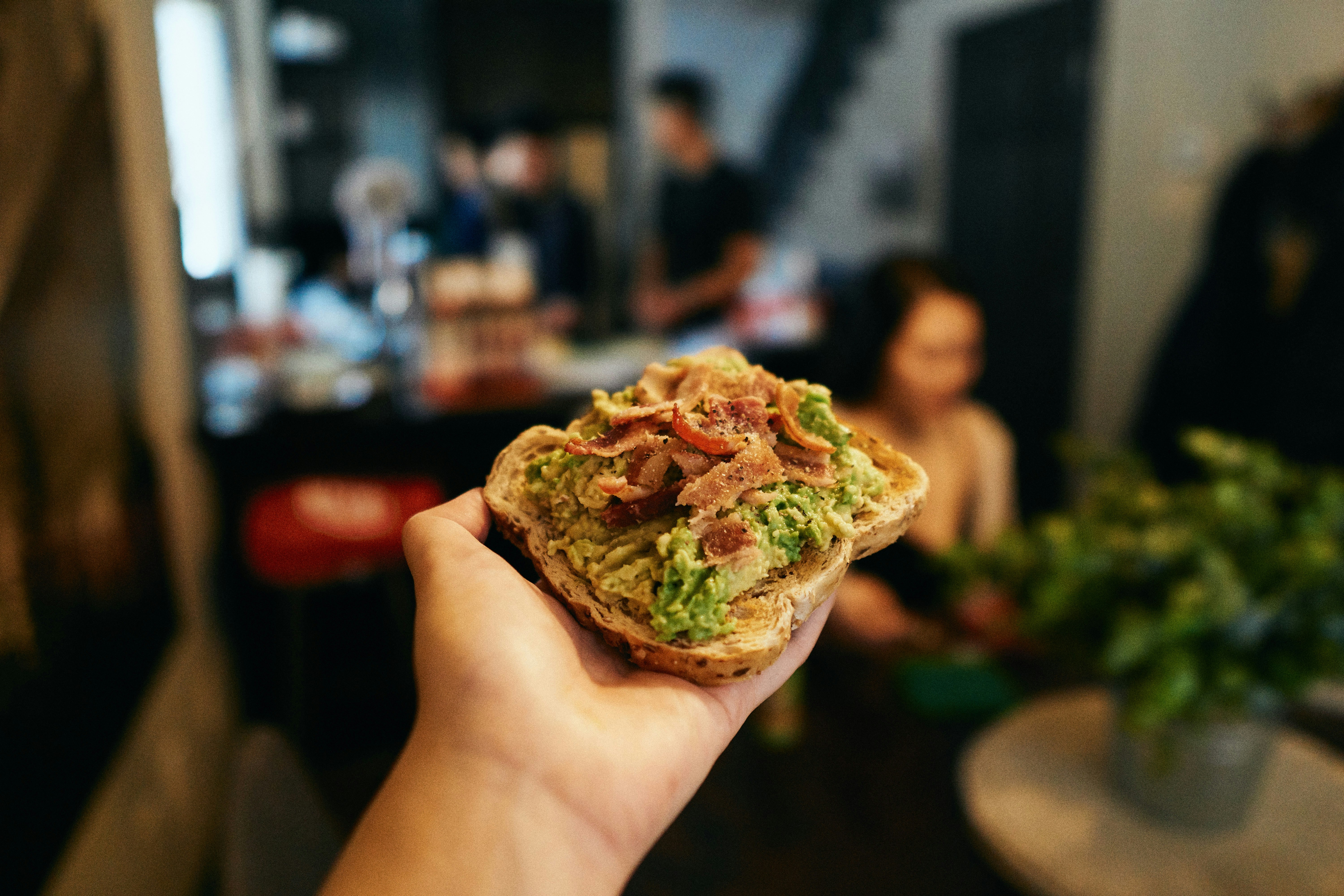 Hand holding a sandwich with fresh ingredients in a cozy indoor setting.