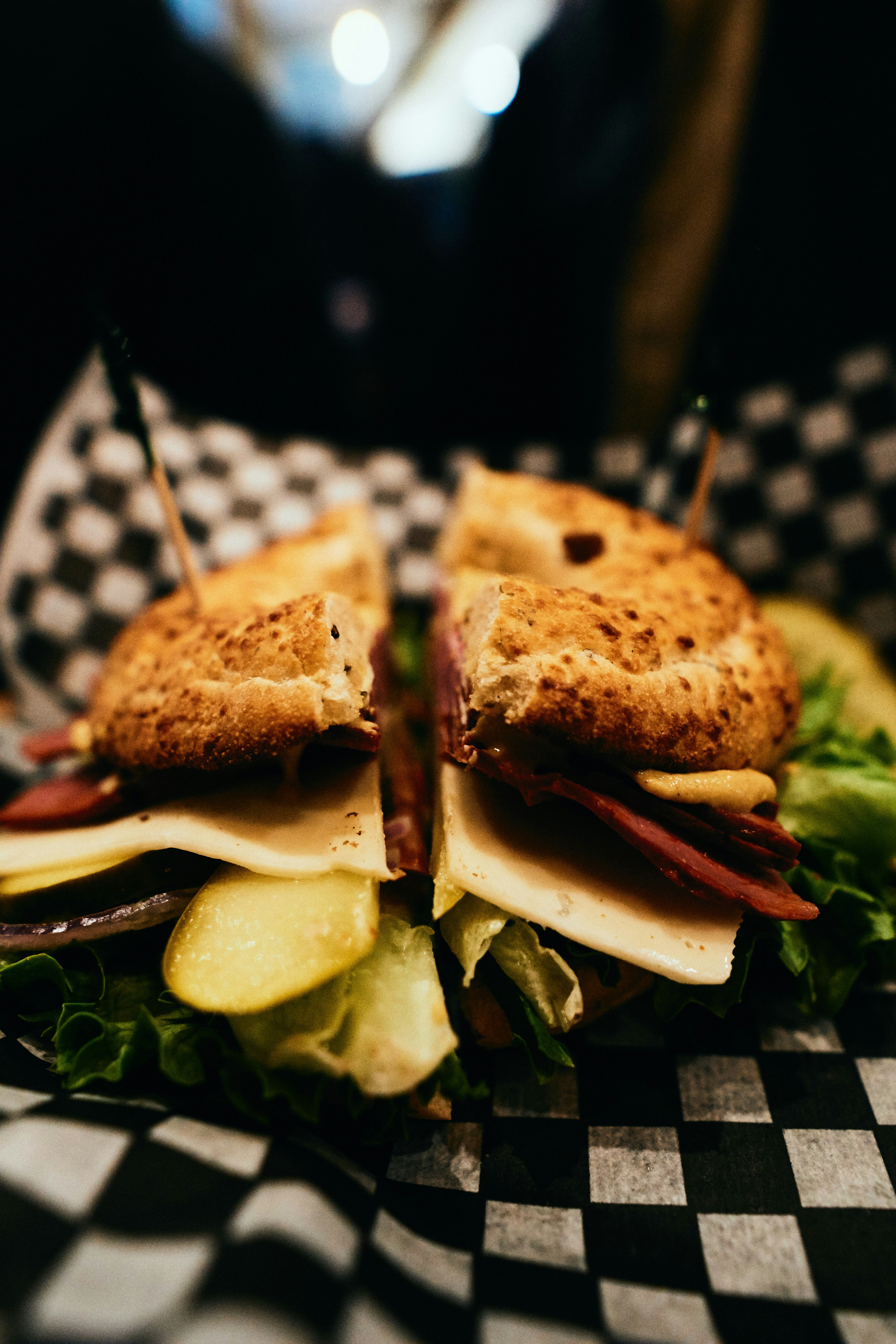 Close-up of a freshly cut artisan sandwich loaded with meats, cheeses, and crisp vegetables, served with pickles on a checkered paper backdrop.