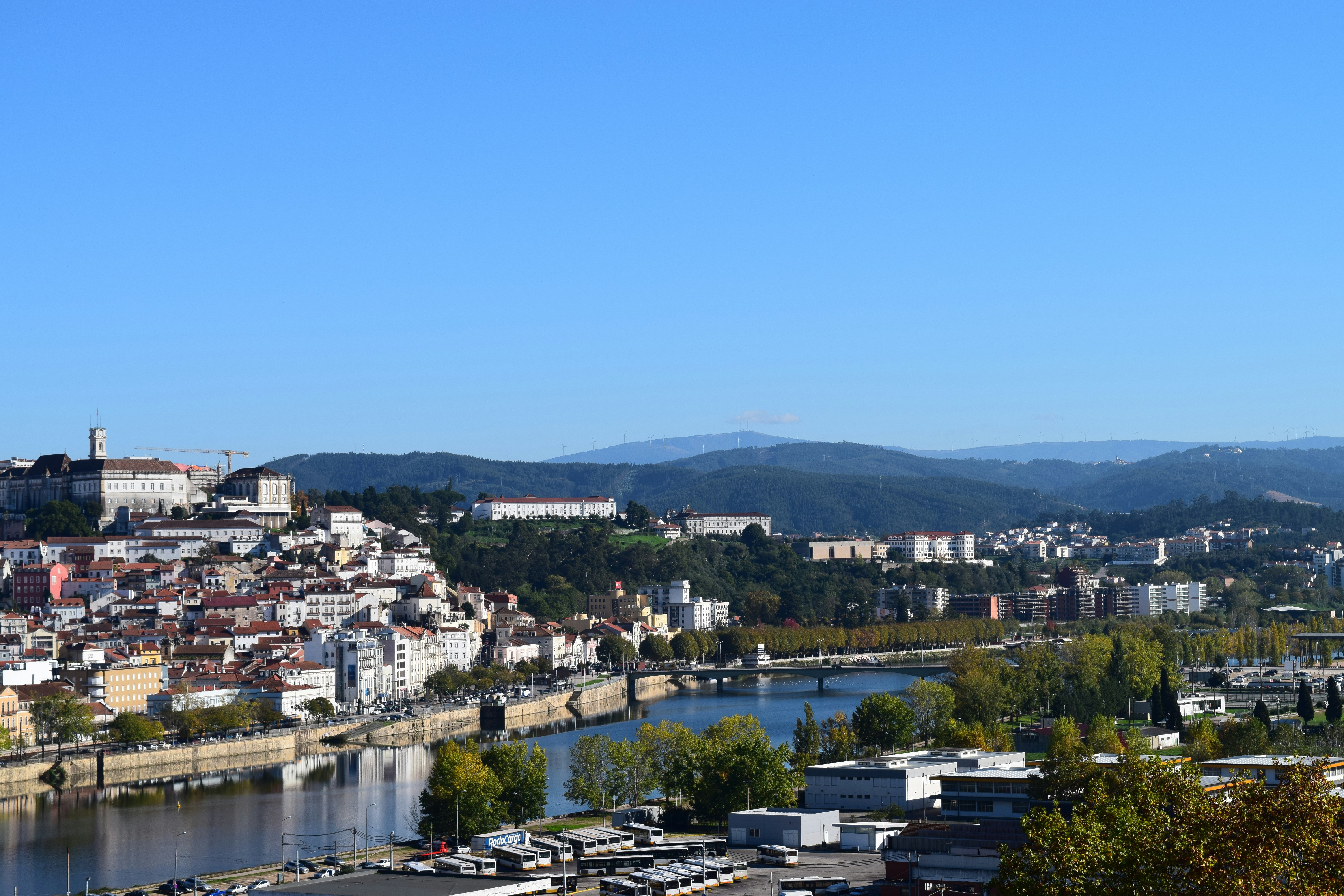 Panoramic view of Coimbra with its historic buildings by the river under a clear blue sky.
