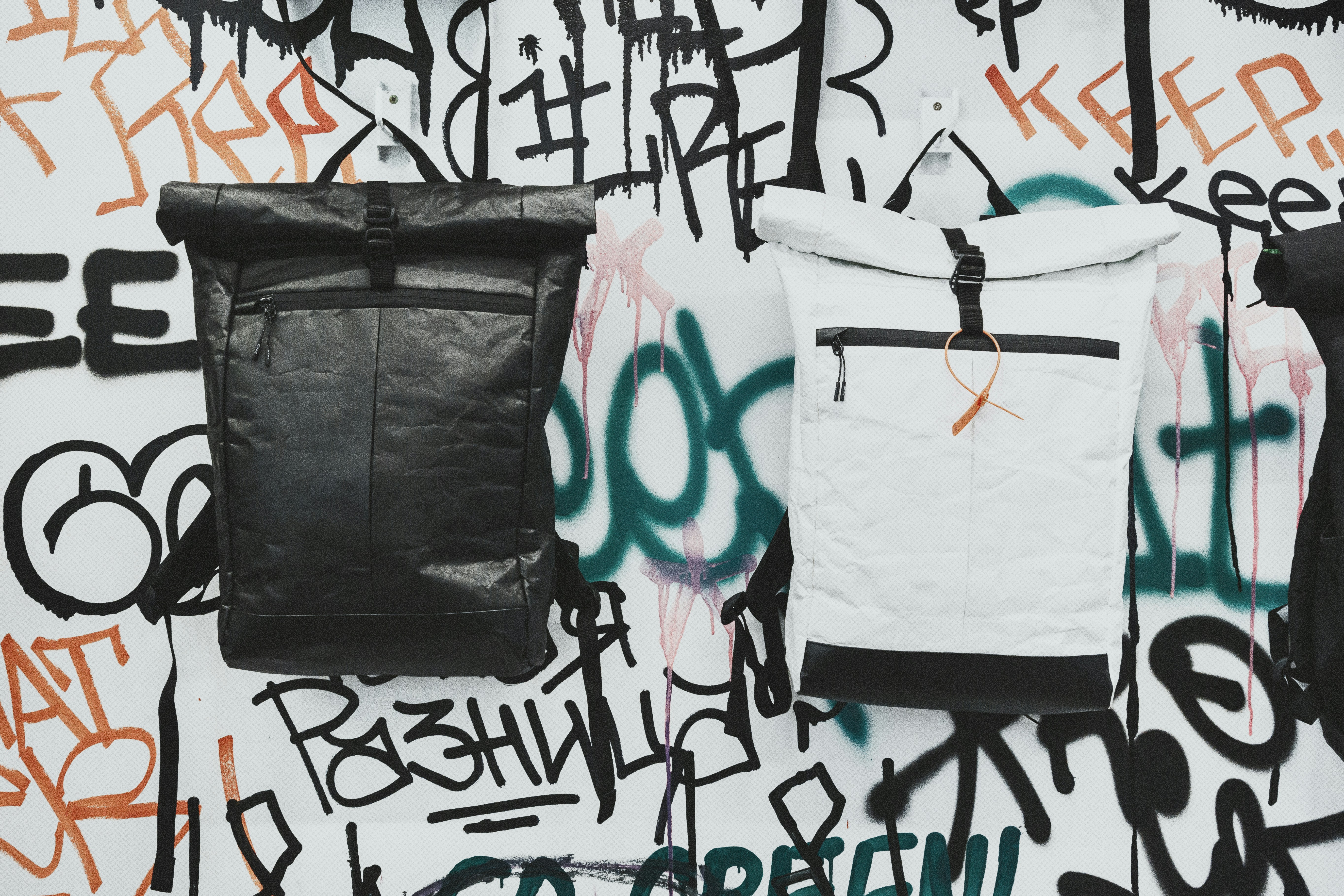 Two backpacks, one black and one white, hang against a graffiti-covered wall, showcasing a blend of urban style and artistry.