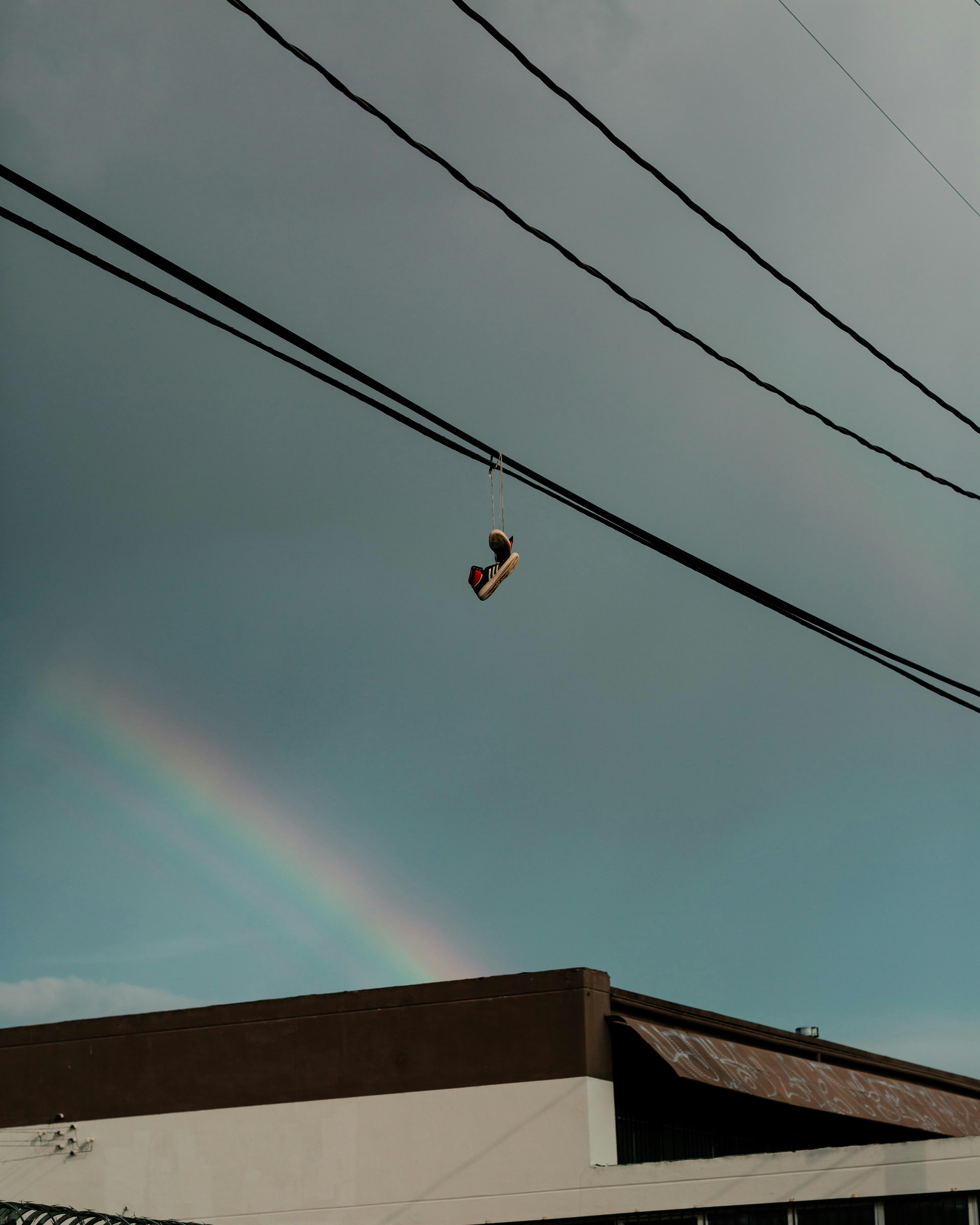 A single sneaker hangs from power lines against a backdrop of moody skies and a faint rainbow. The scene captures an urban moment of unexpected whimsy.
