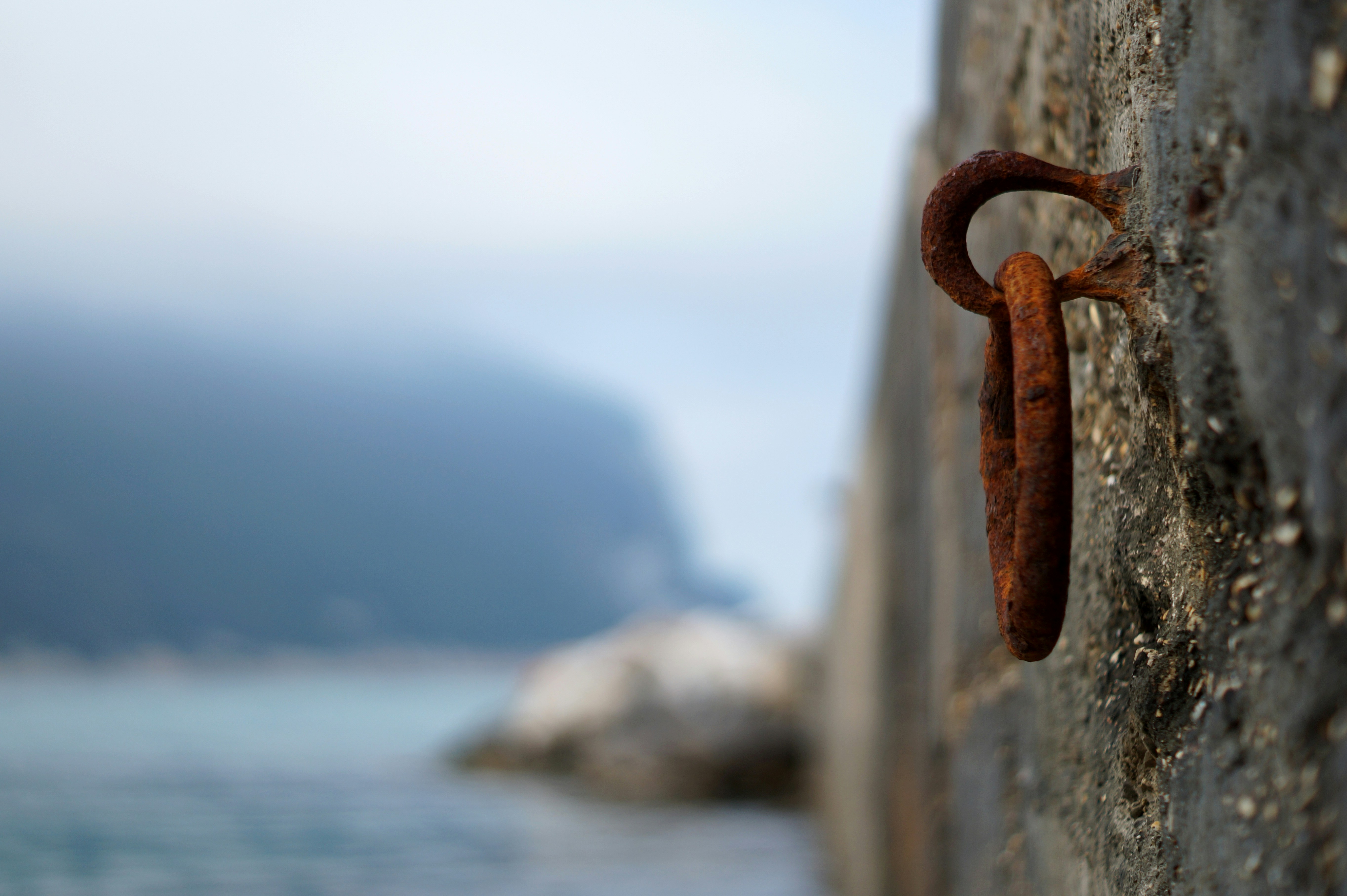 A rusted metal hook embedded in a coastal rock wall with a misty sea and mountains in the background.