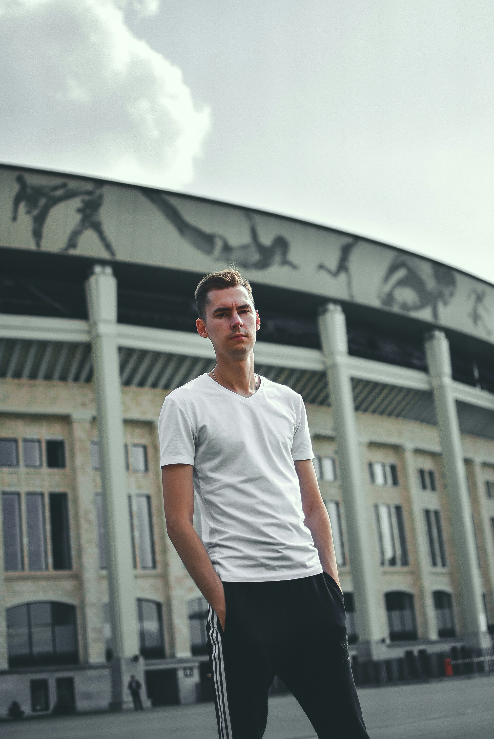 Young man in a white t-shirt and black track pants stands confidently in front of a grand sports stadium, showcasing a blend of urban style and architectural beauty.