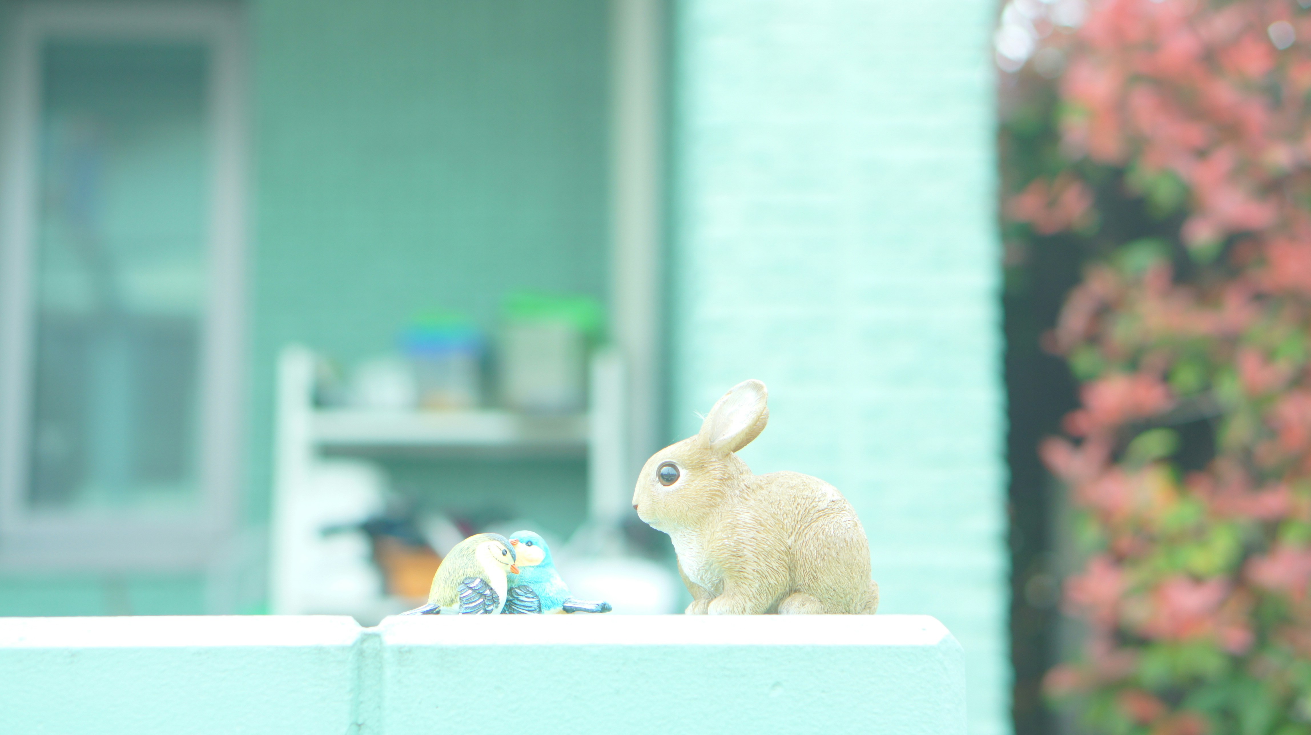 A charming scene featuring a rabbit figurine and a colorful bird perched on a ledge, set against a soft, blurred background of greenery and household items.