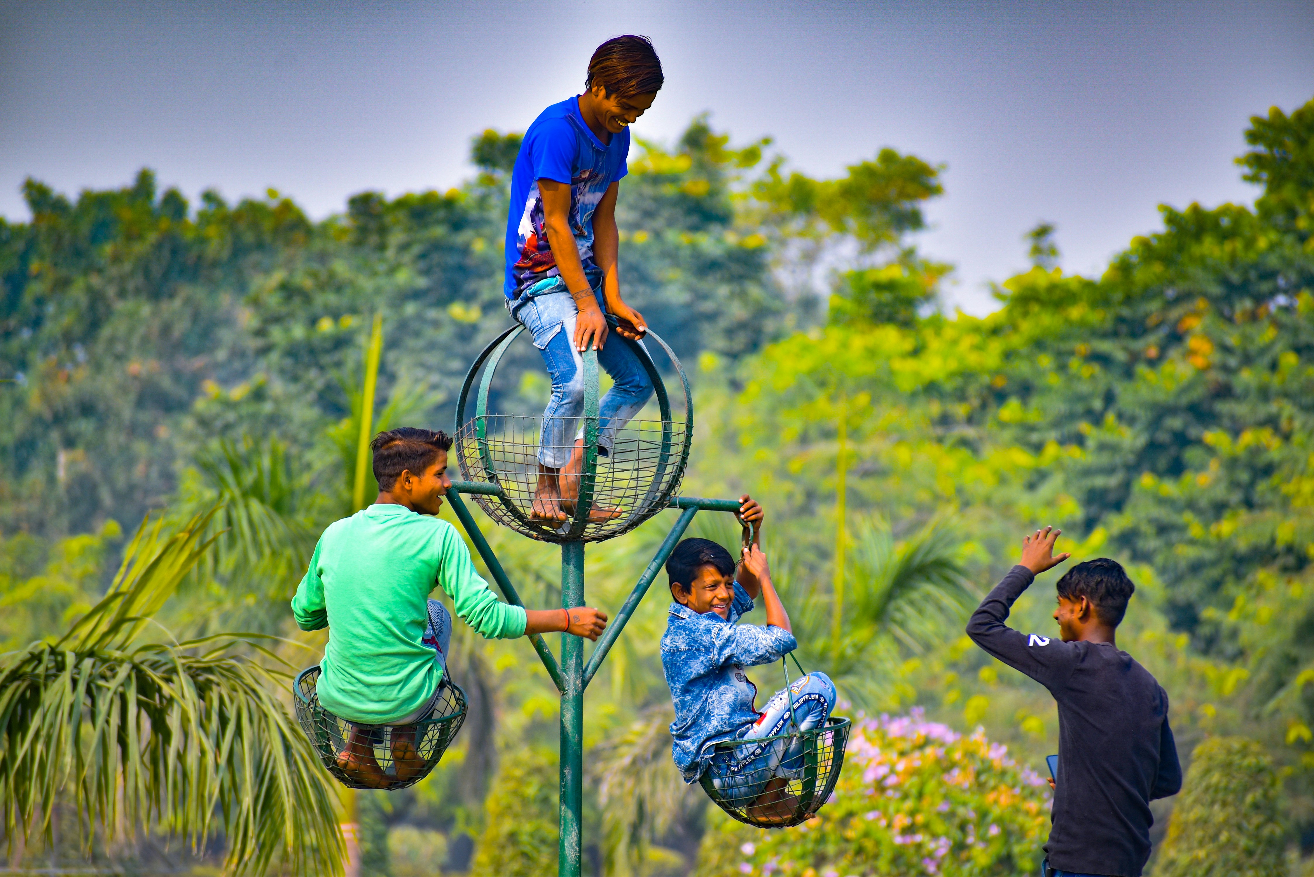 Group of youths climbing a circular playground structure surrounded by lush greenery.
