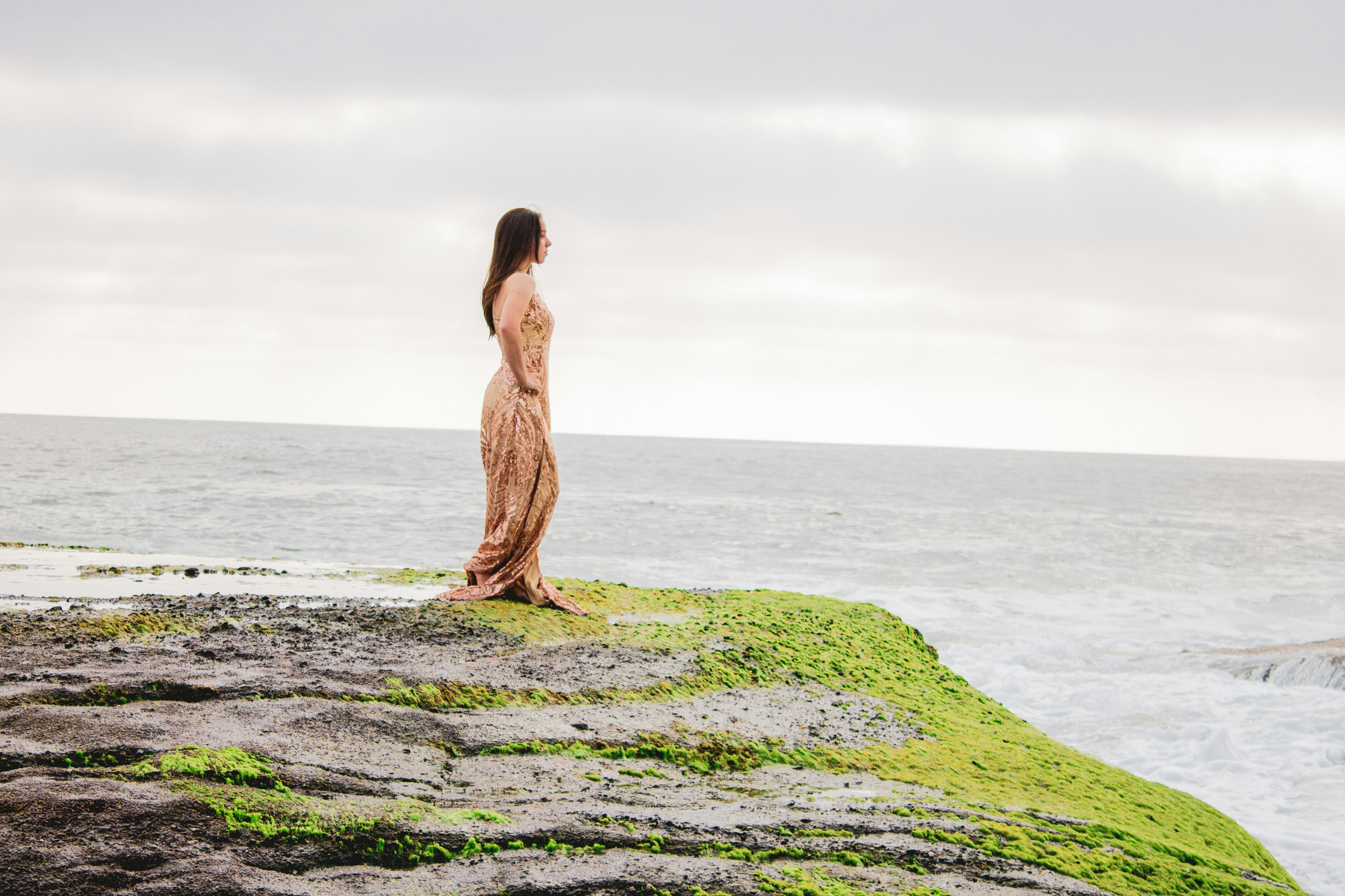 Solitary Horizon: Woman in a flowing dress on mossy rock jutting into the sea