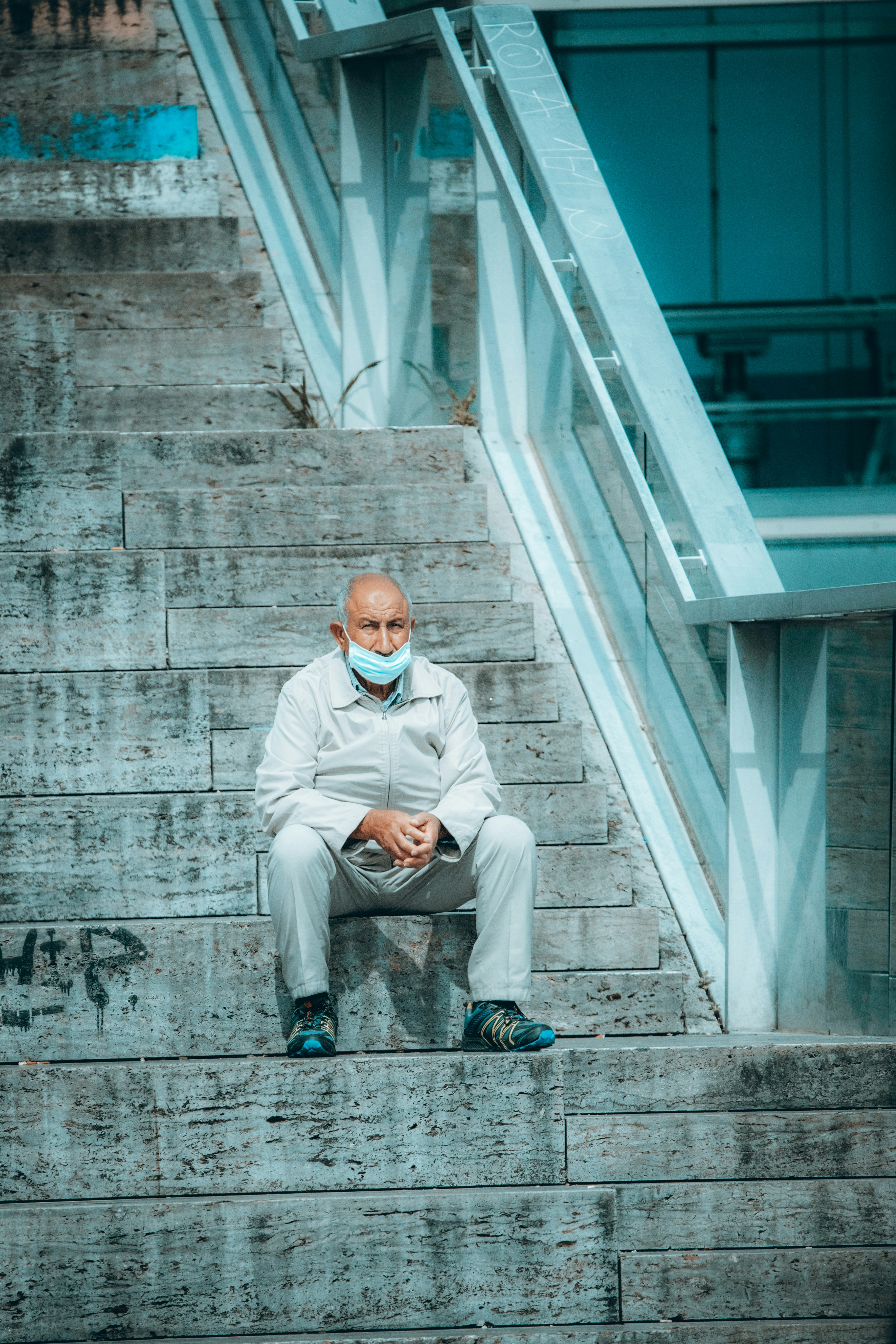 An elderly man seated on concrete steps, wearing a mask, surrounded by modern architecture. The scene captures a moment of reflection in an urban setting.