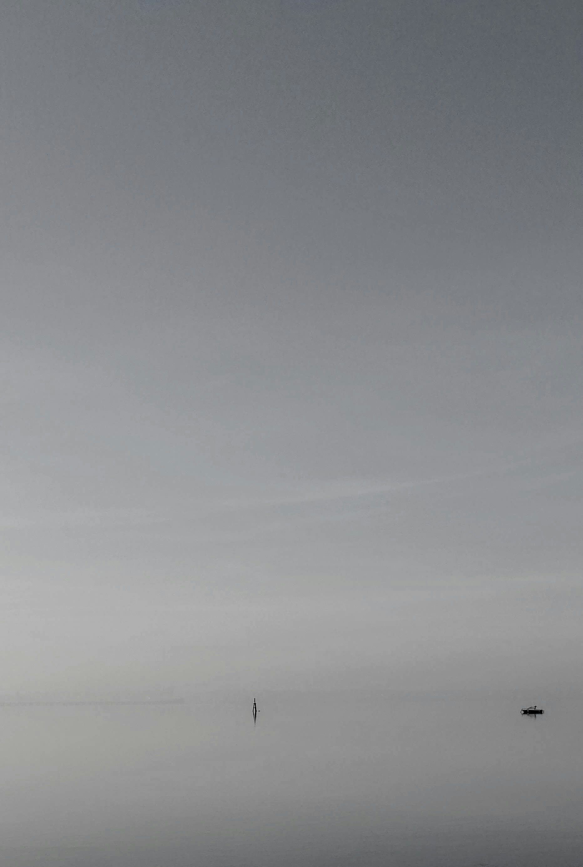 A solitary pole stands in the serene expanse of a calm body of water, mirrored by the soft, muted sky above.