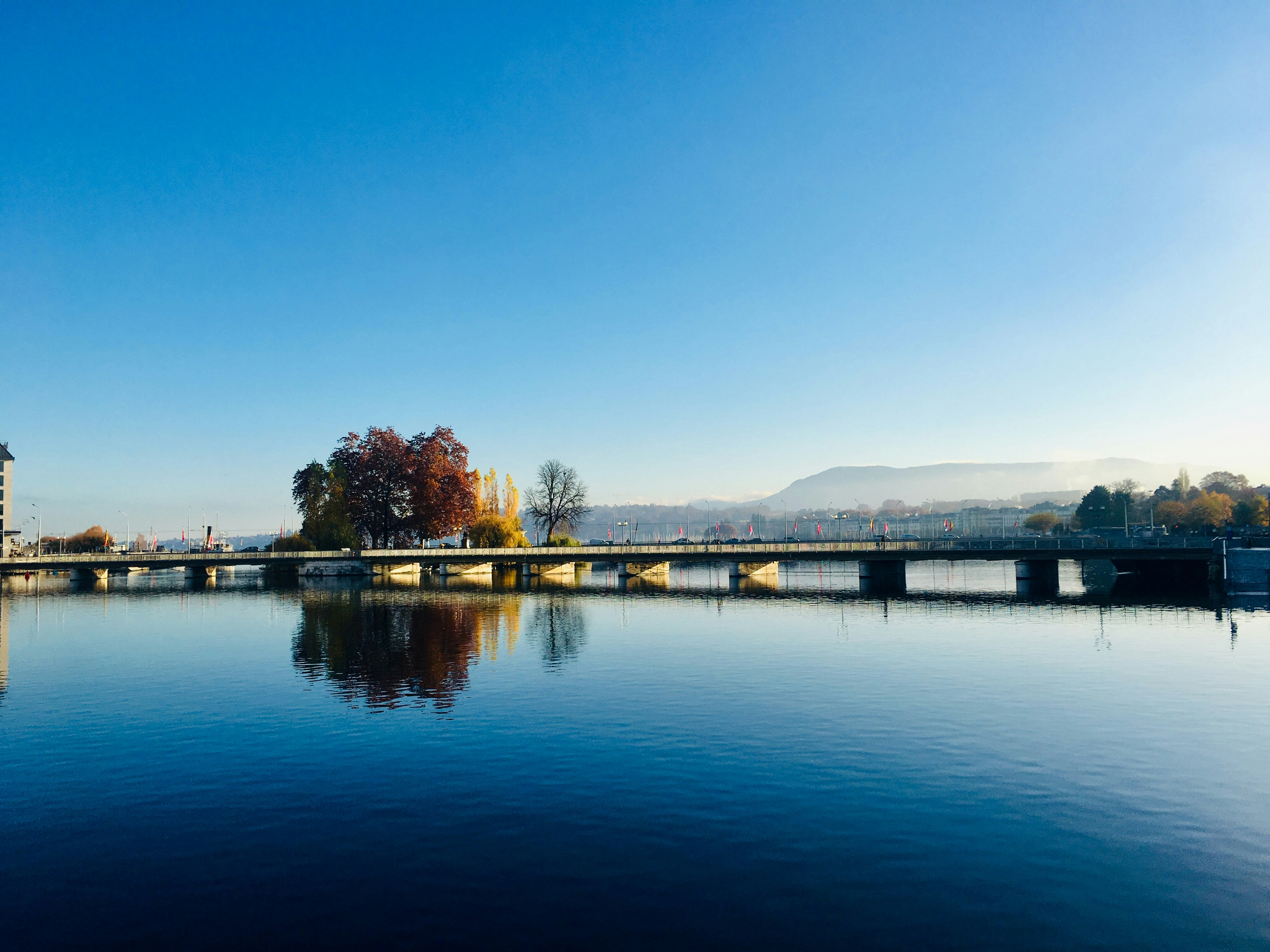 Calm water reflects a distant bridge under a clear blue sky.