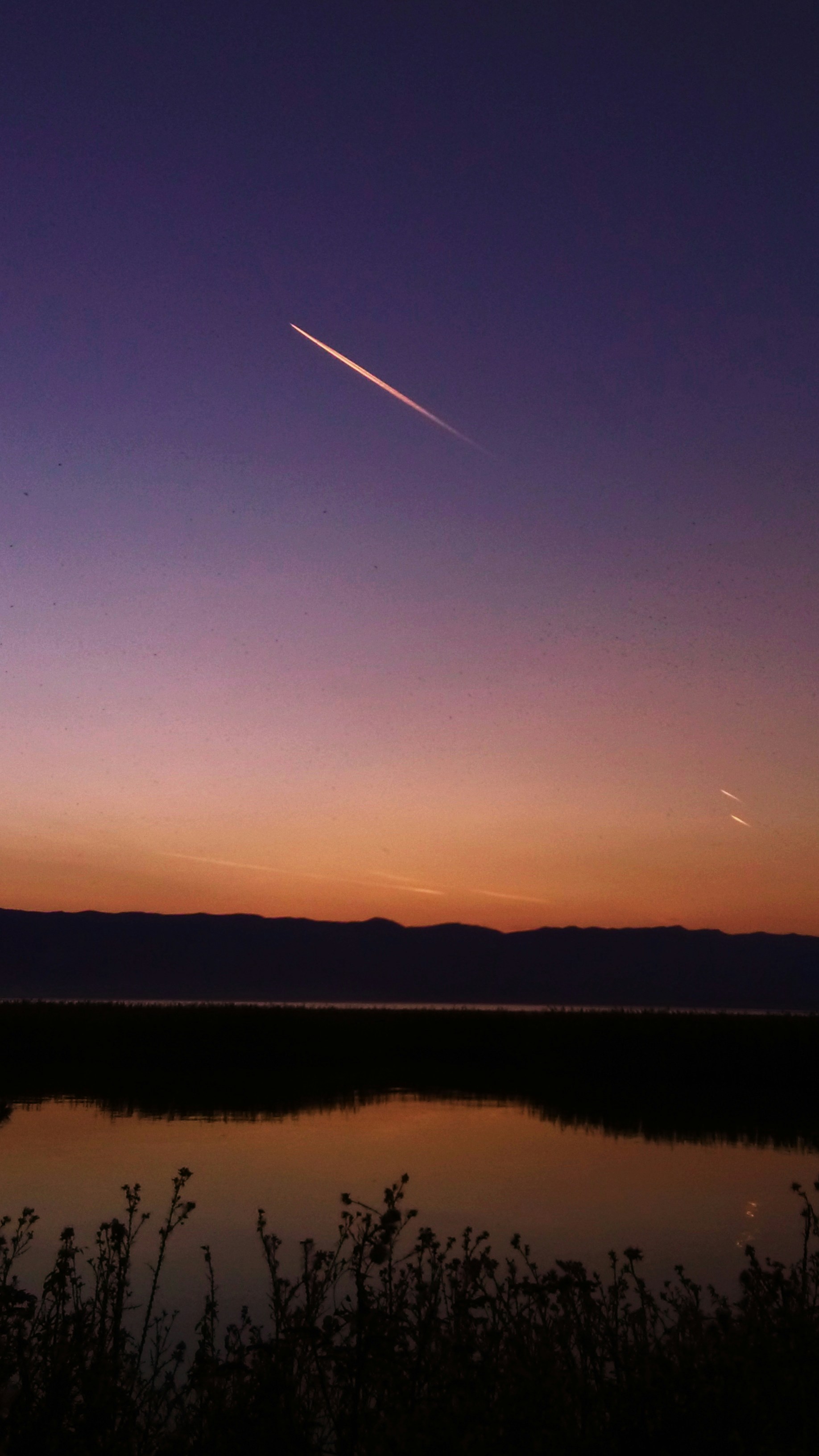 A serene lakeside view at dusk, with a faint contrail streaking across the twilight sky and reflections shimmering on the water's surface.