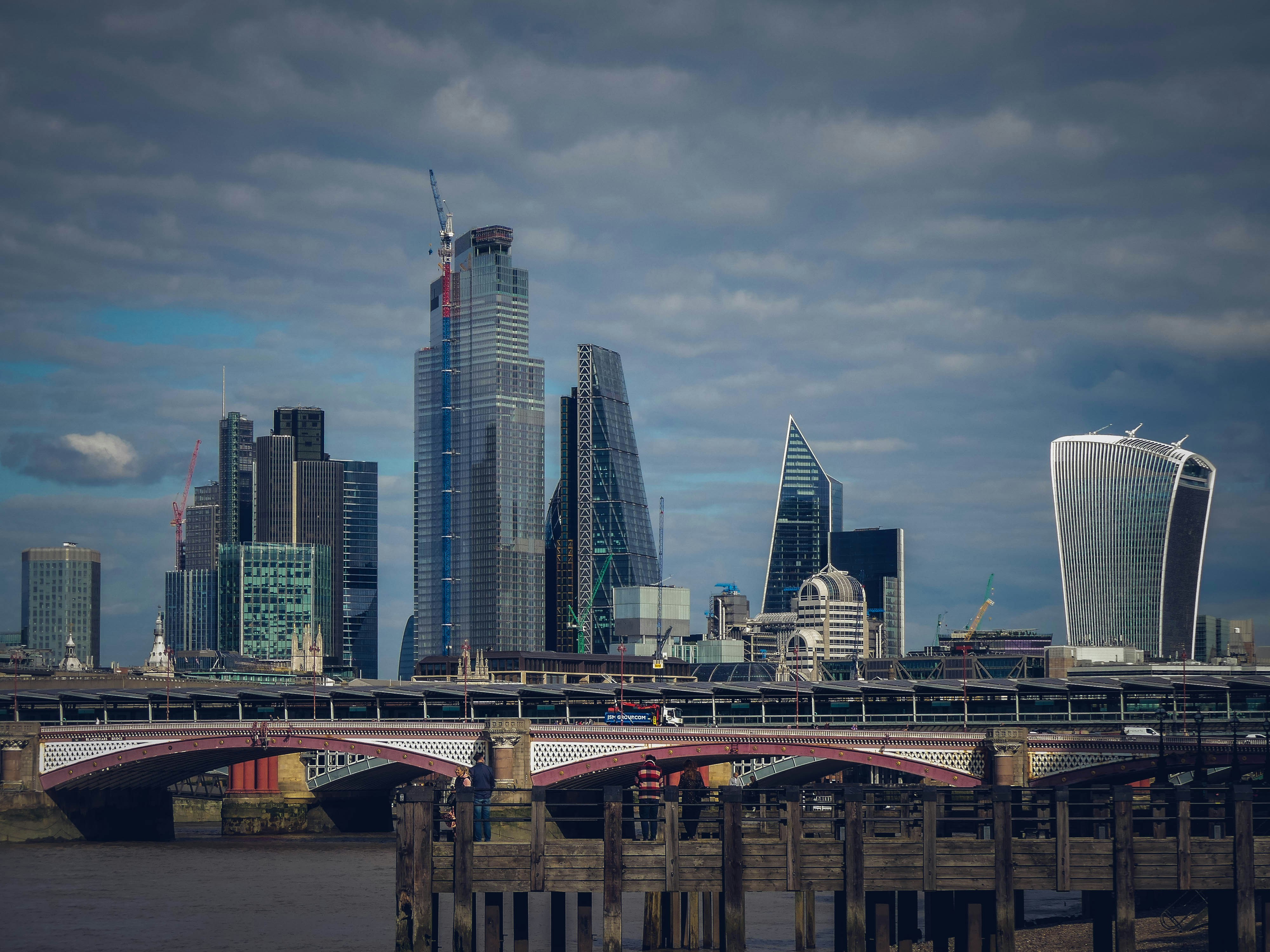 A vibrant cityscape showcasing London's iconic skyscrapers juxtaposed against a cloudy sky, with a bridge in the foreground. The scene highlights the architectural diversity of the metropolis.