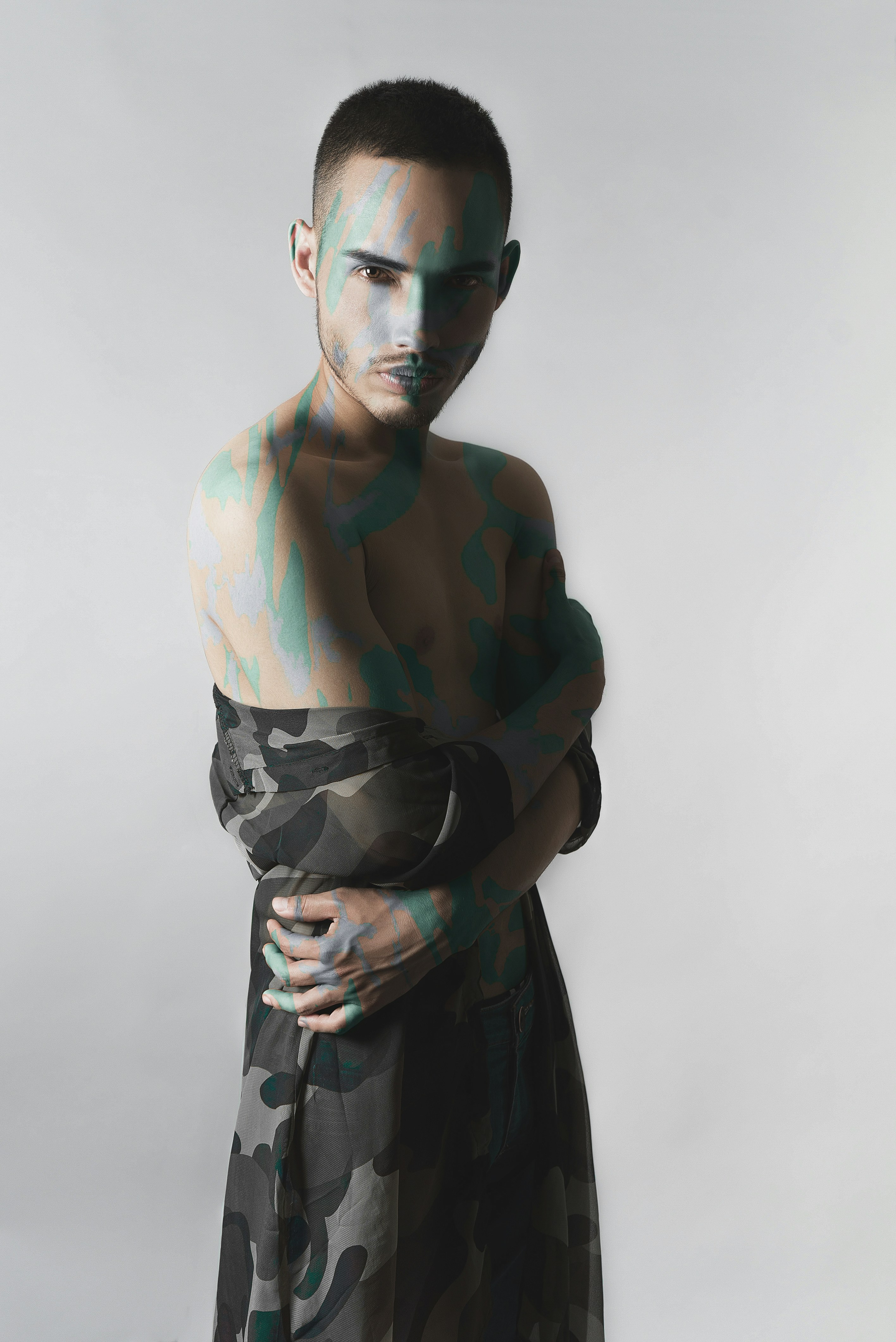 A male figure with painted patterns on his face and arms, draped in a camouflage garment, poses against a minimalist background.