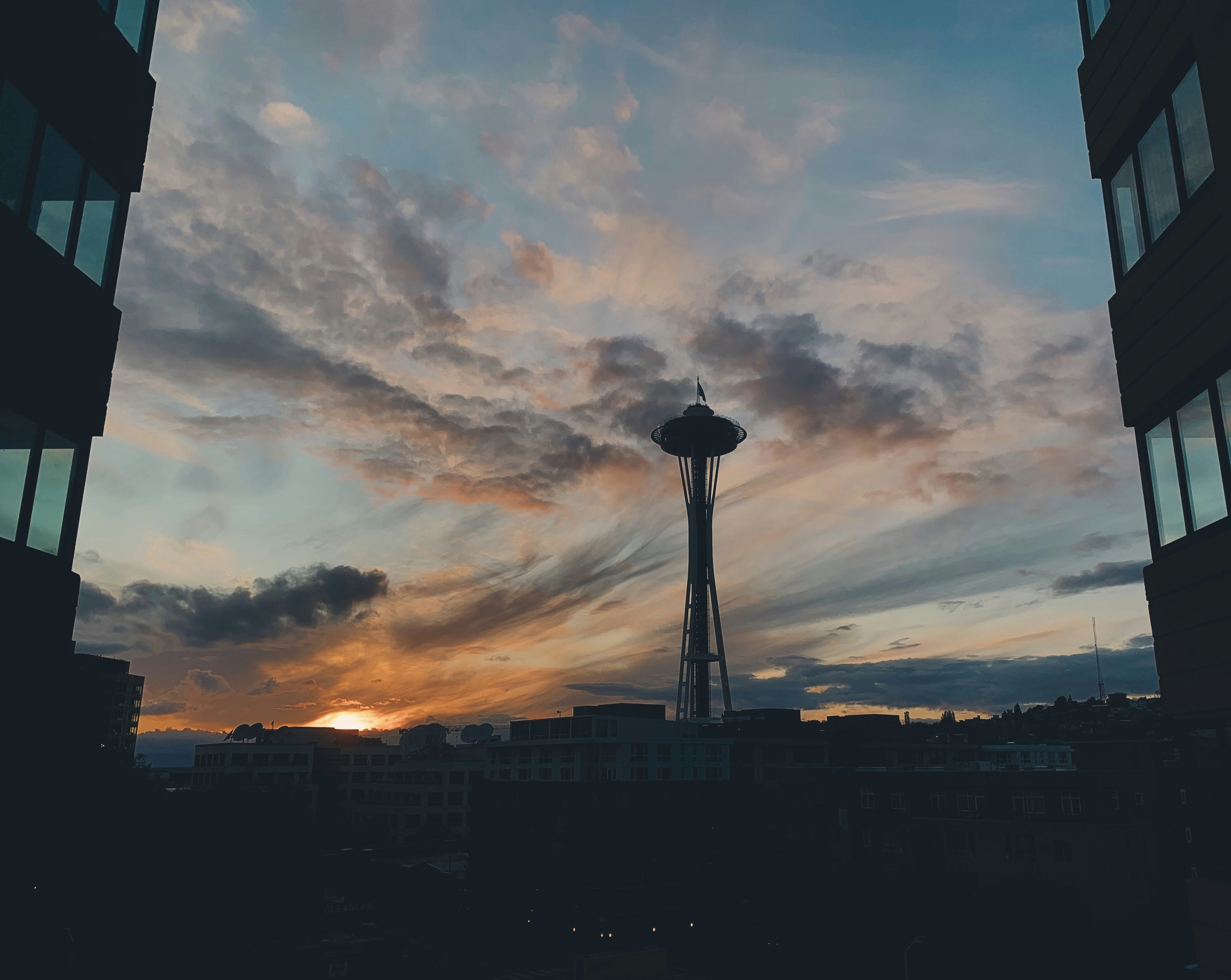 Silhouetted Space Needle against a vibrant sunset sky, framed by modern buildings. The clouds create a dynamic backdrop.