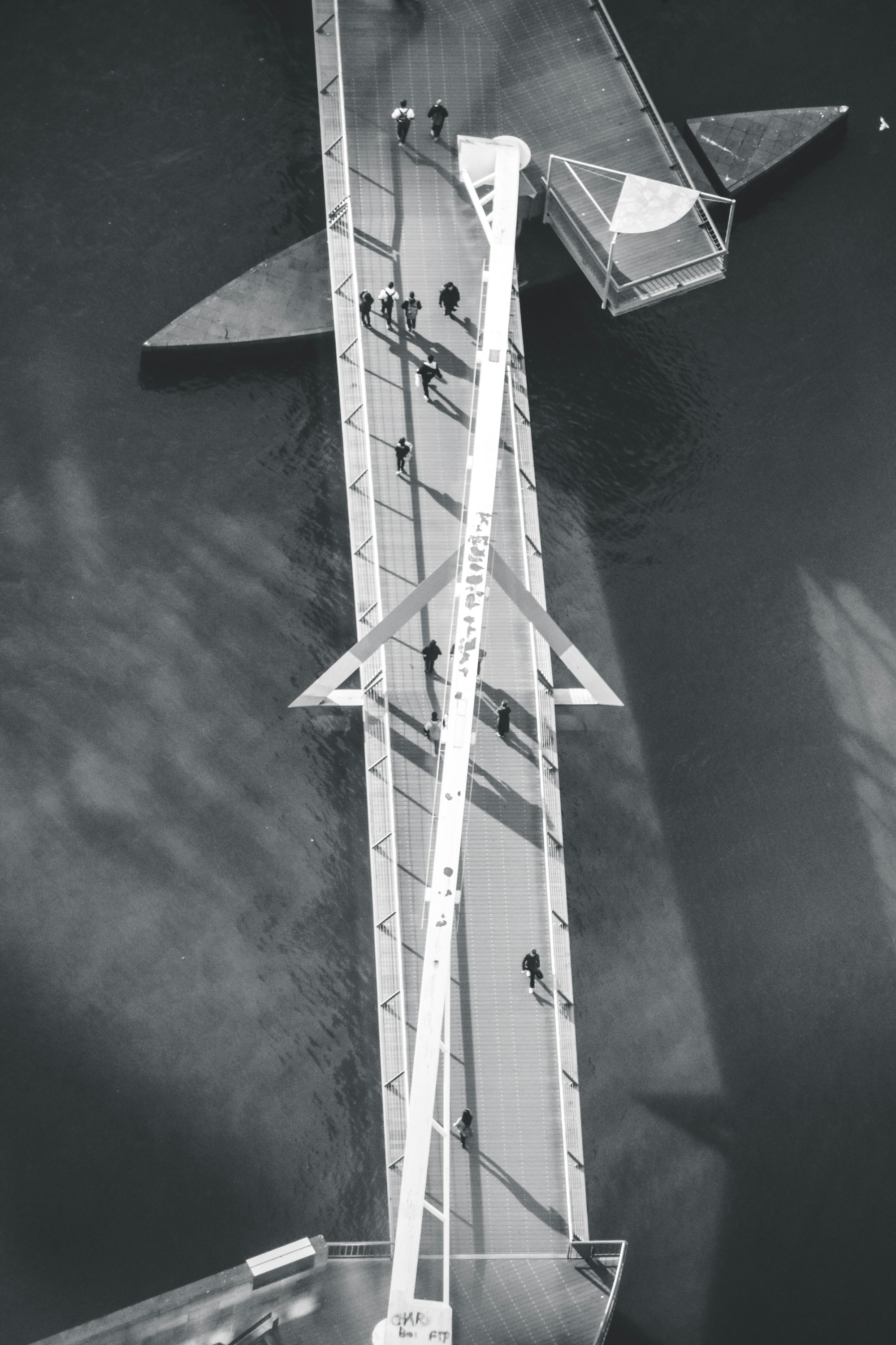 Aerial view of a modern pedestrian bridge with individuals walking across, showcasing geometric lines and reflections on the water below.