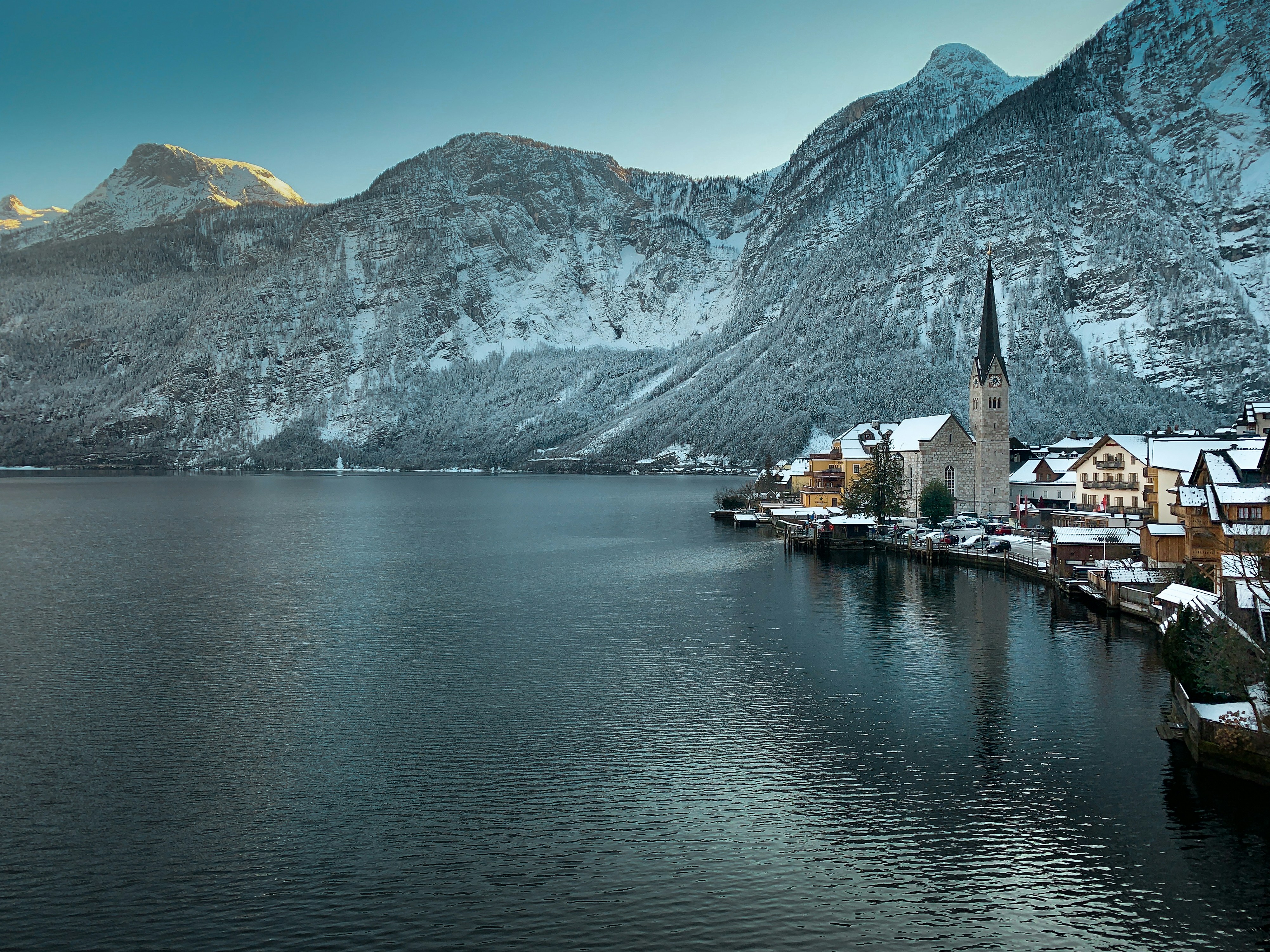 Snow-covered village nestled by a tranquil lake with majestic mountains in the background.