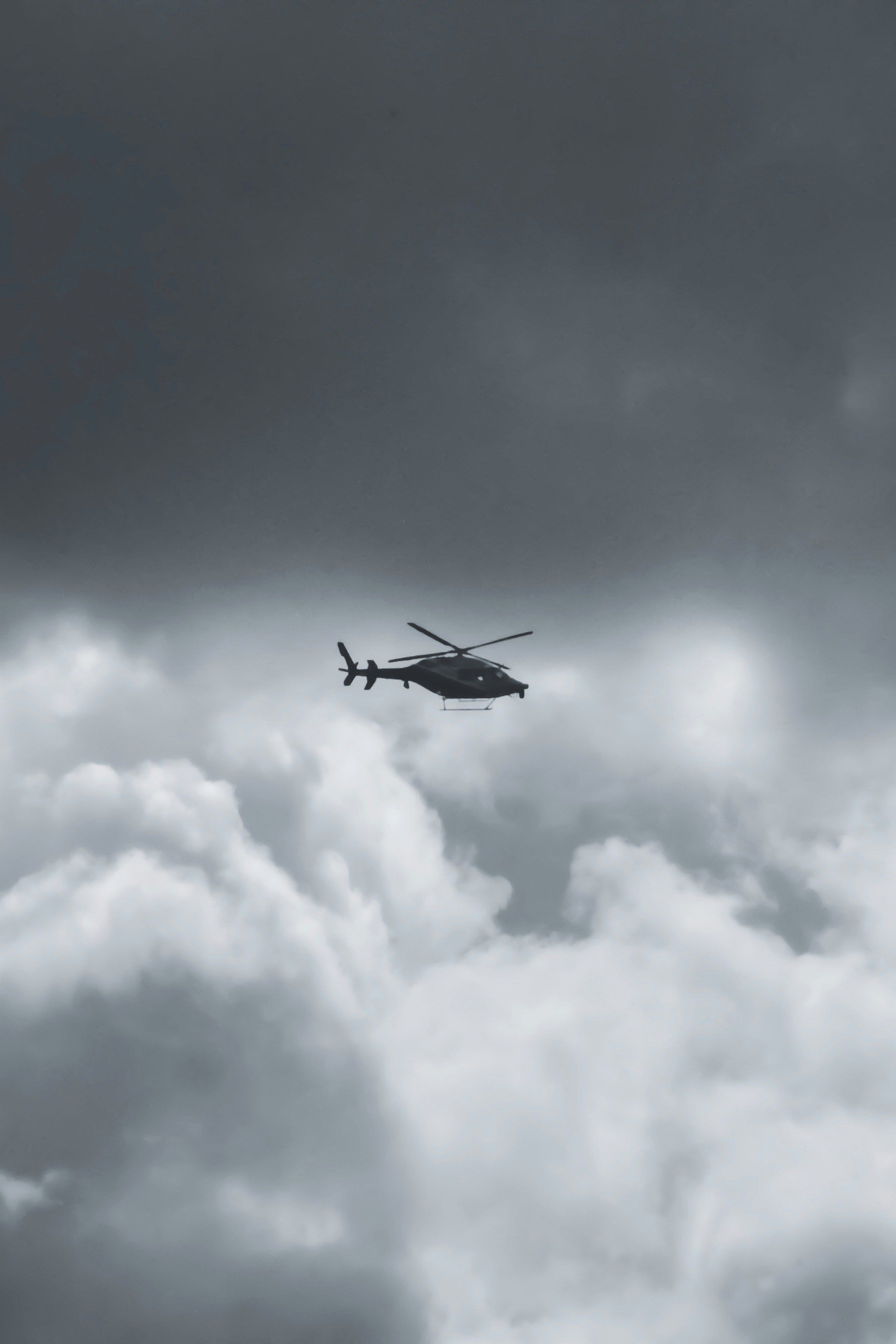 A lone helicopter silhouette drifts through heavy storm clouds in a moody aerial photograph. The scene emphasizes scale and weather-driven tension.