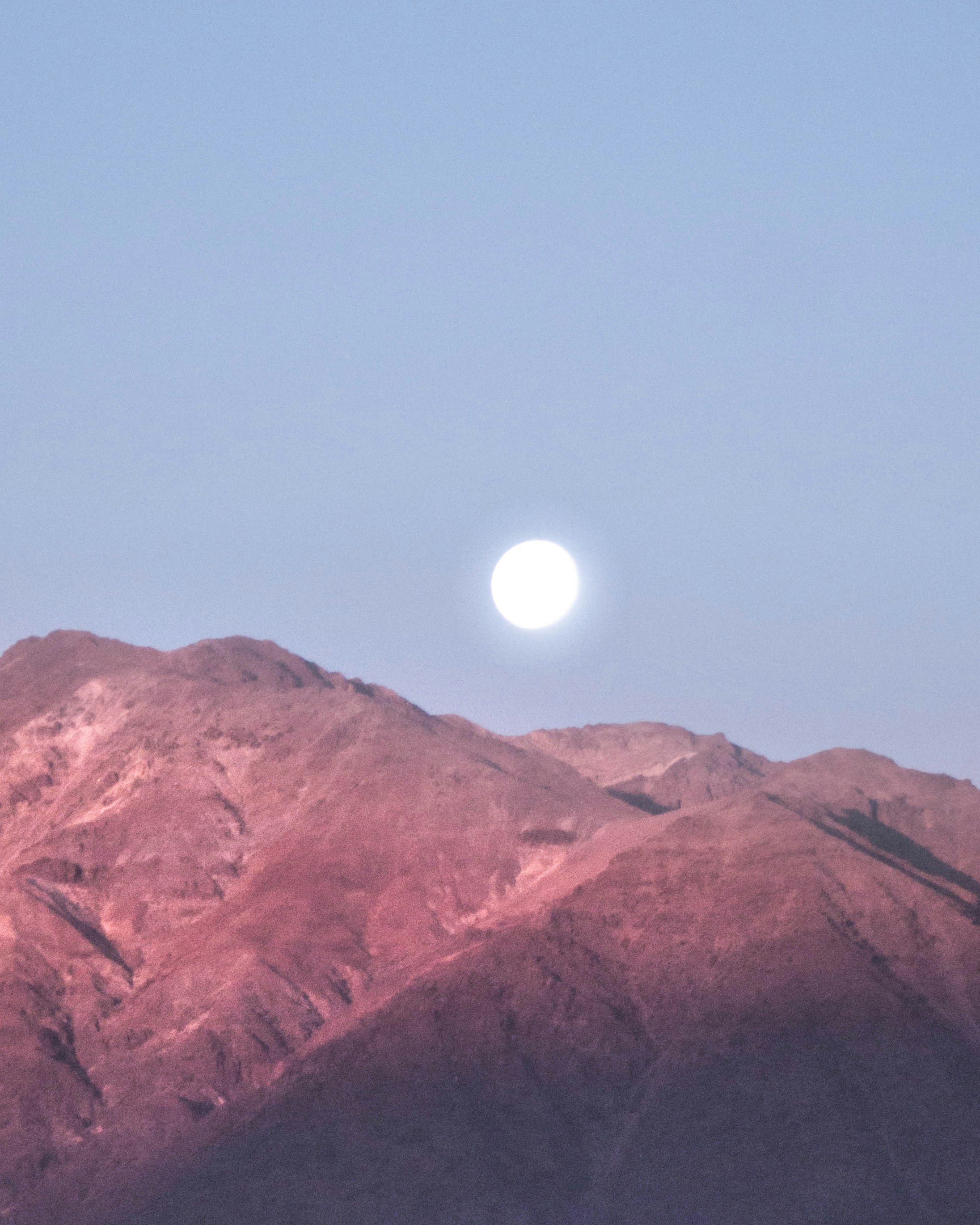Full moon rising above mountains bathed in soft pink light against a clear sky.