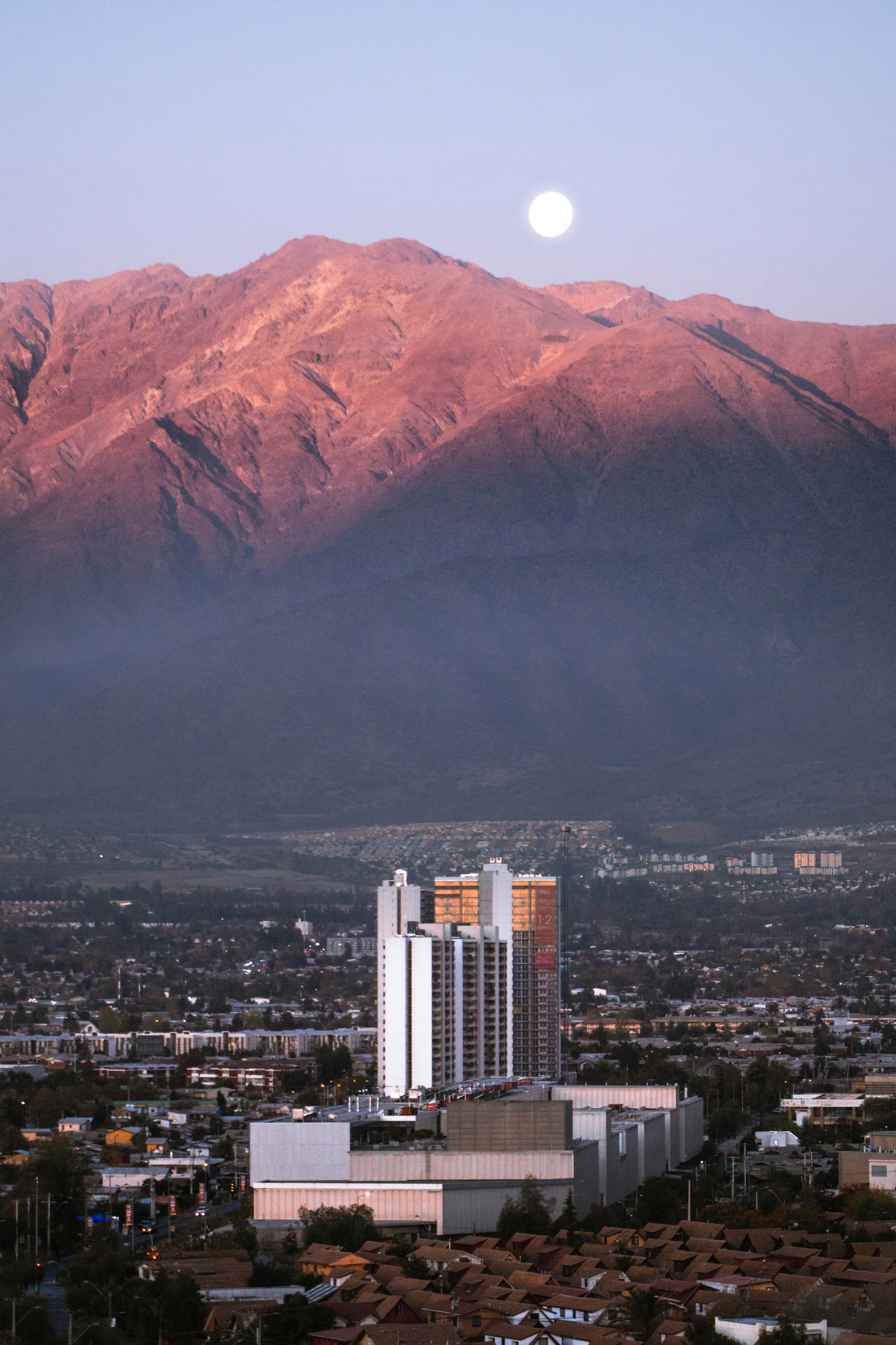 Moon rising above a cityscape with tall buildings, framed by mountains in the evening light.